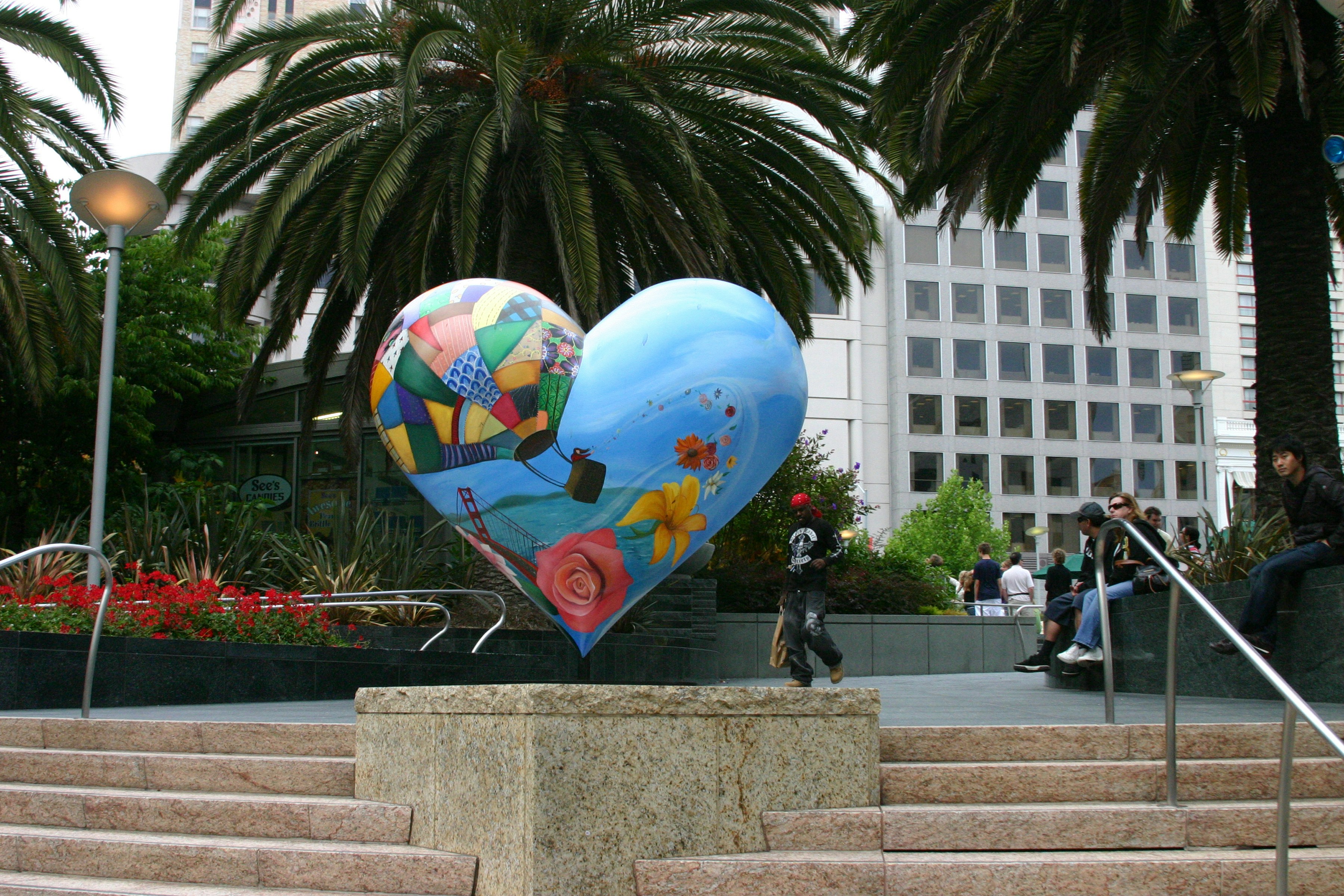 Vibrantly painted heart sculpture surrounded by palm trees and urban greenery, serving as a public art installation. The scene captures a blend of nature and artistic expression.