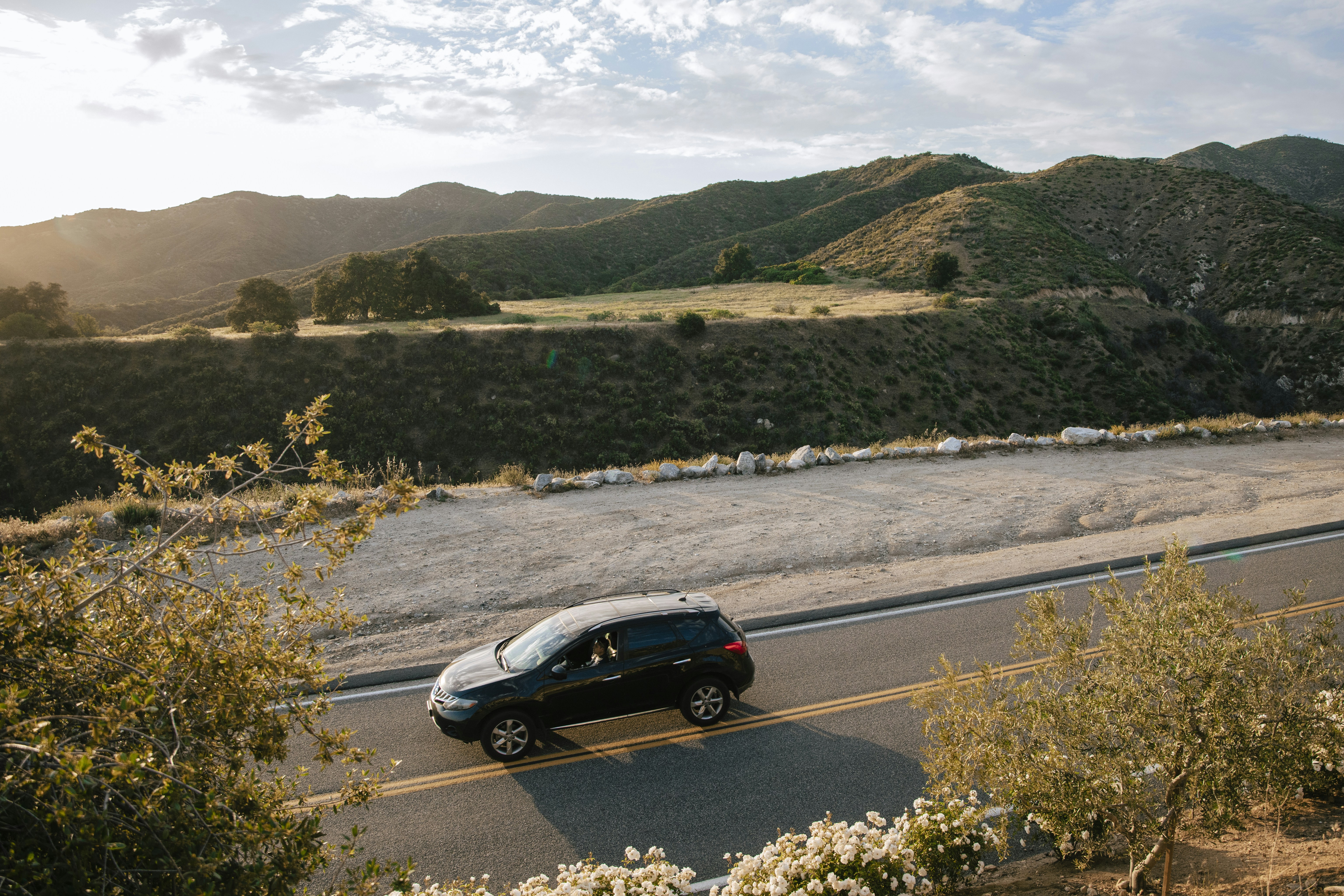 A black SUV drives along a winding road surrounded by lush hills and a golden sunset glow.