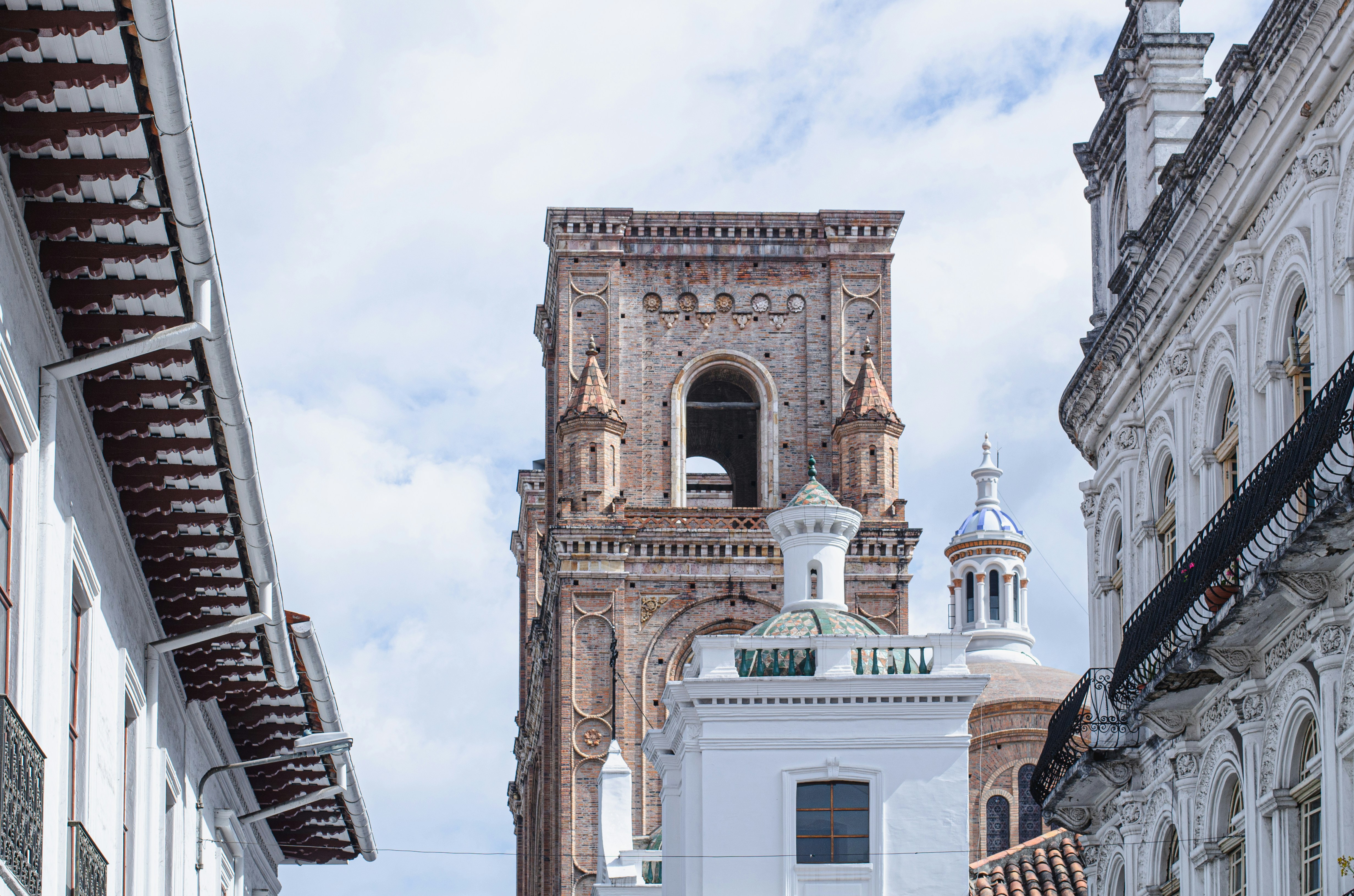 Tall brick tower rises between ornate white buildings under a partly cloudy sky.