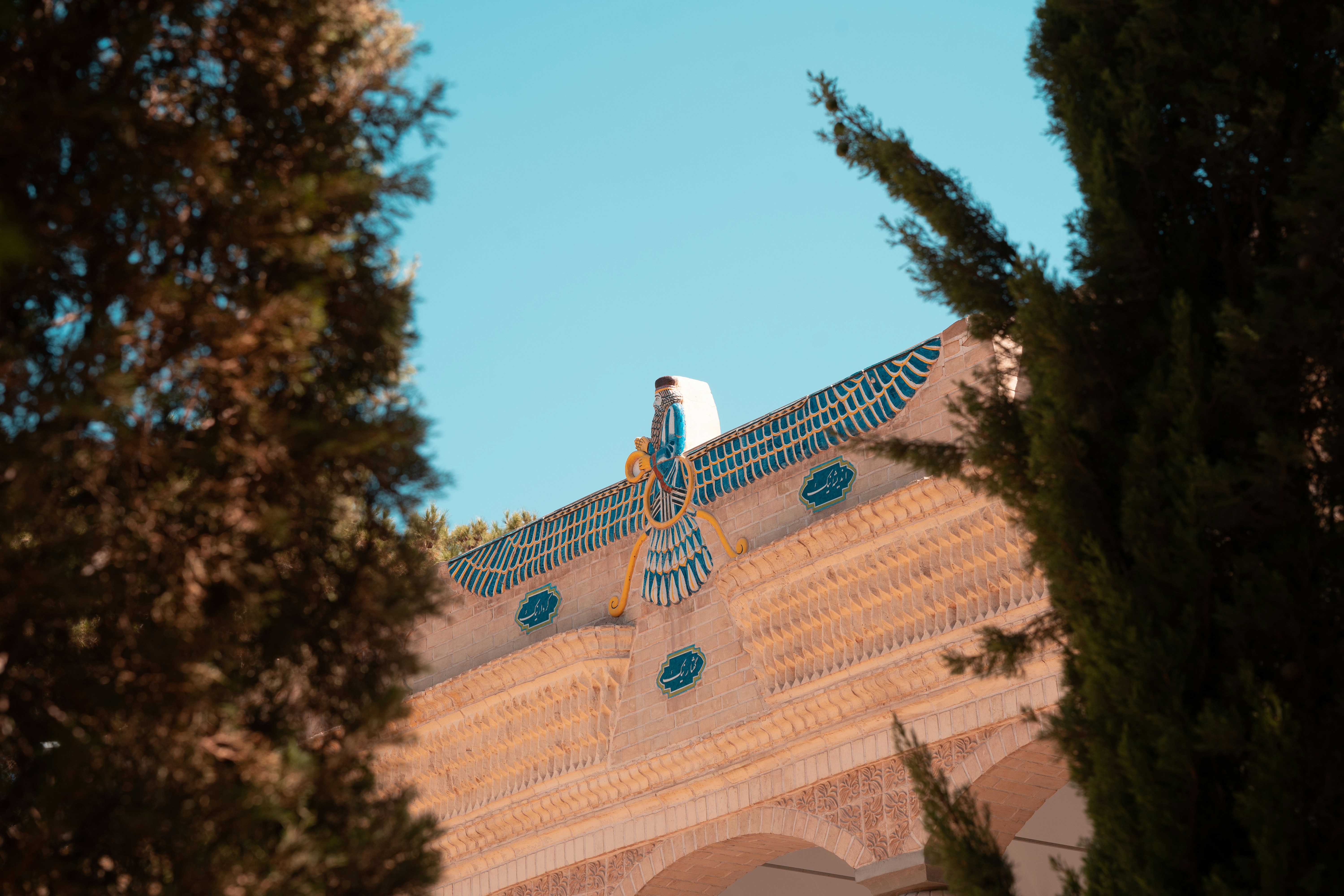 Intricate architectural detail featuring a winged figure atop a structure, framed by lush greenery. The design reflects historical artistry and cultural significance.
