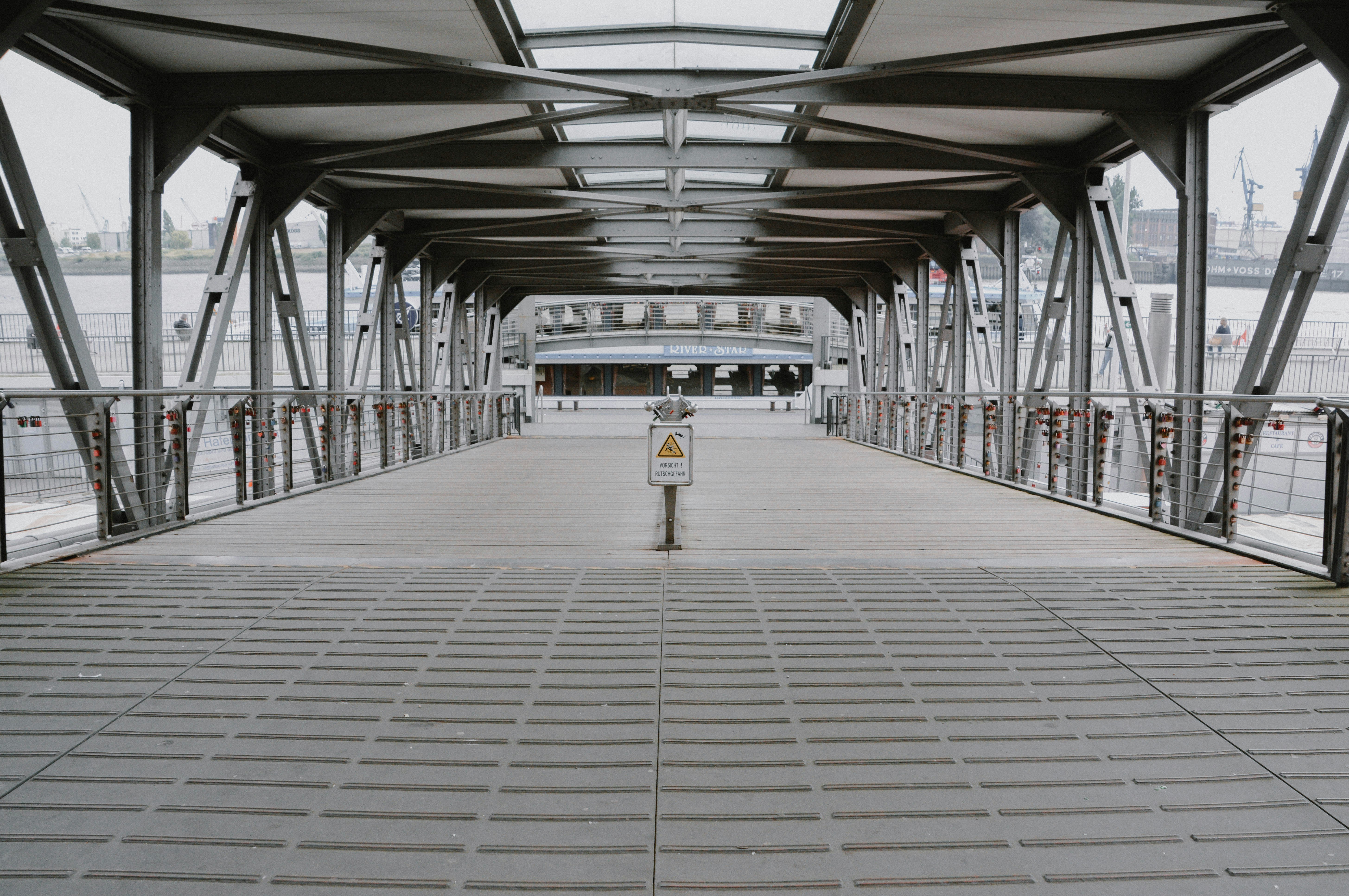A modern pedestrian bridge featuring a symmetrical design with metal beams and a warning sign at the center. The scene captures the essence of contemporary urban architecture.