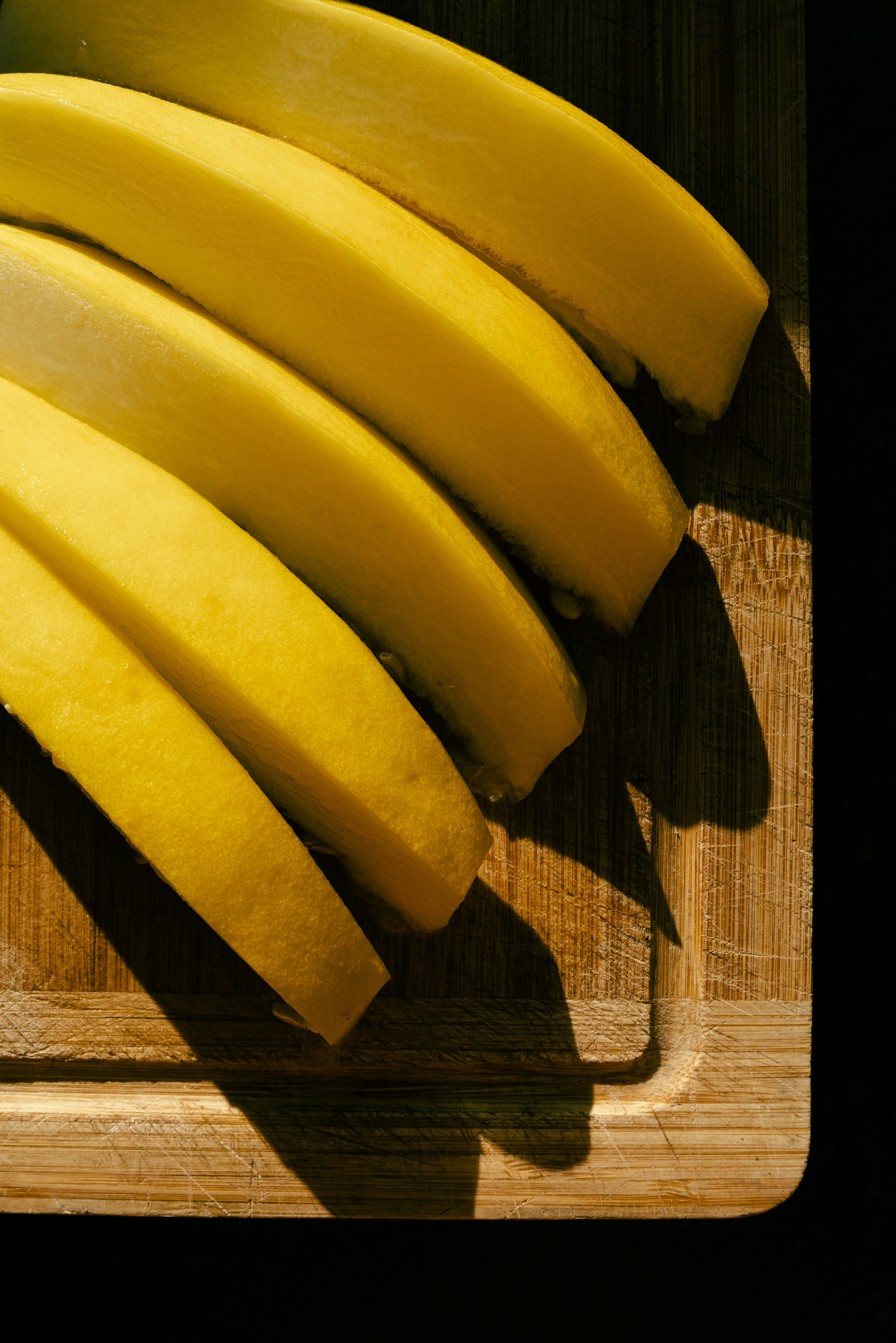 Bright yellow bananas arranged in curved overlapping slices on a wooden cutting board. Side lighting creates bold shadows and emphasizes texture.