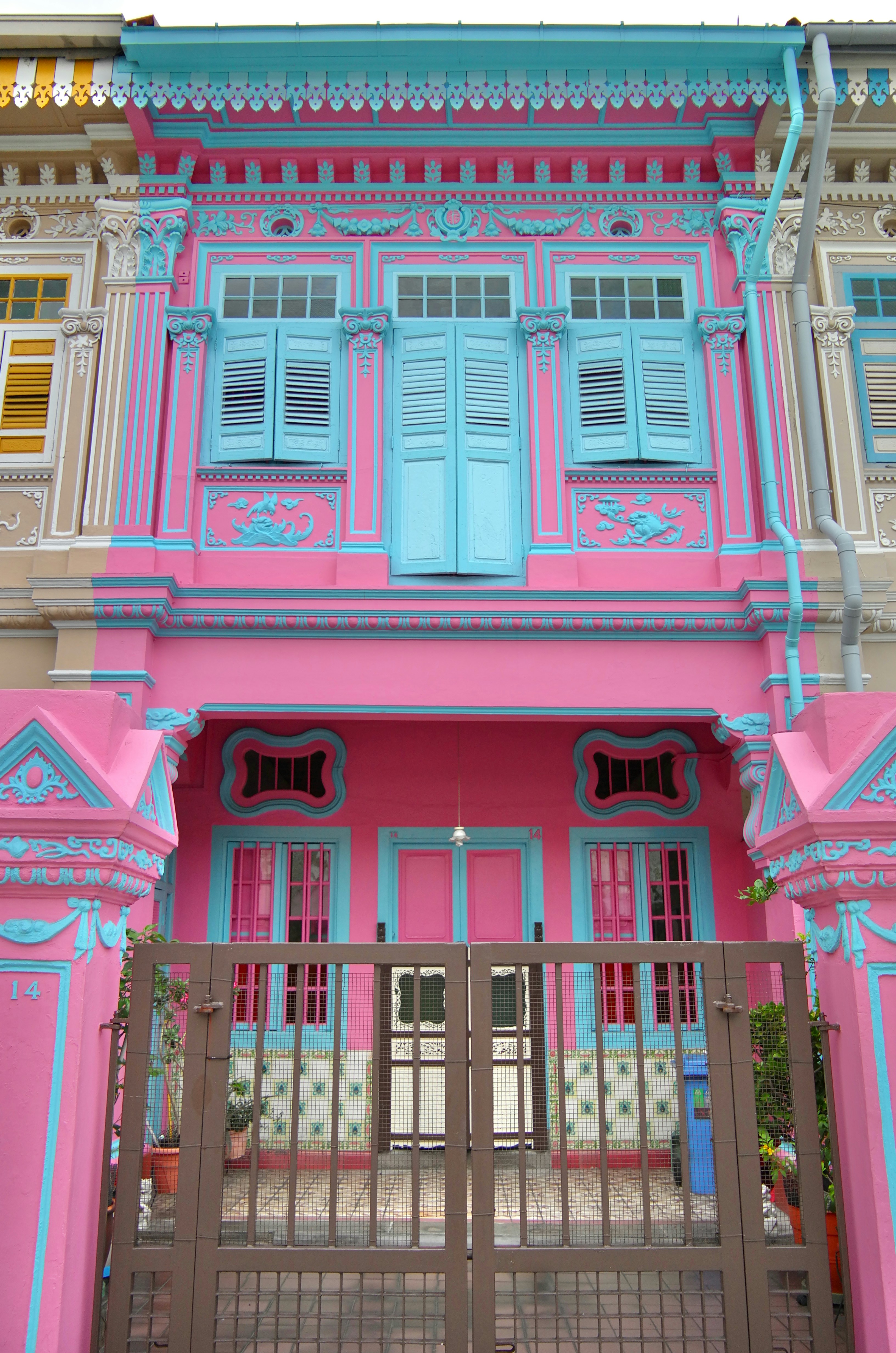 Colorful townhouse façade in pink and blue with ornate trim and a central blue-shuttered window. A brown gate frames the ground-floor entrance, highlighting architectural whimsy.