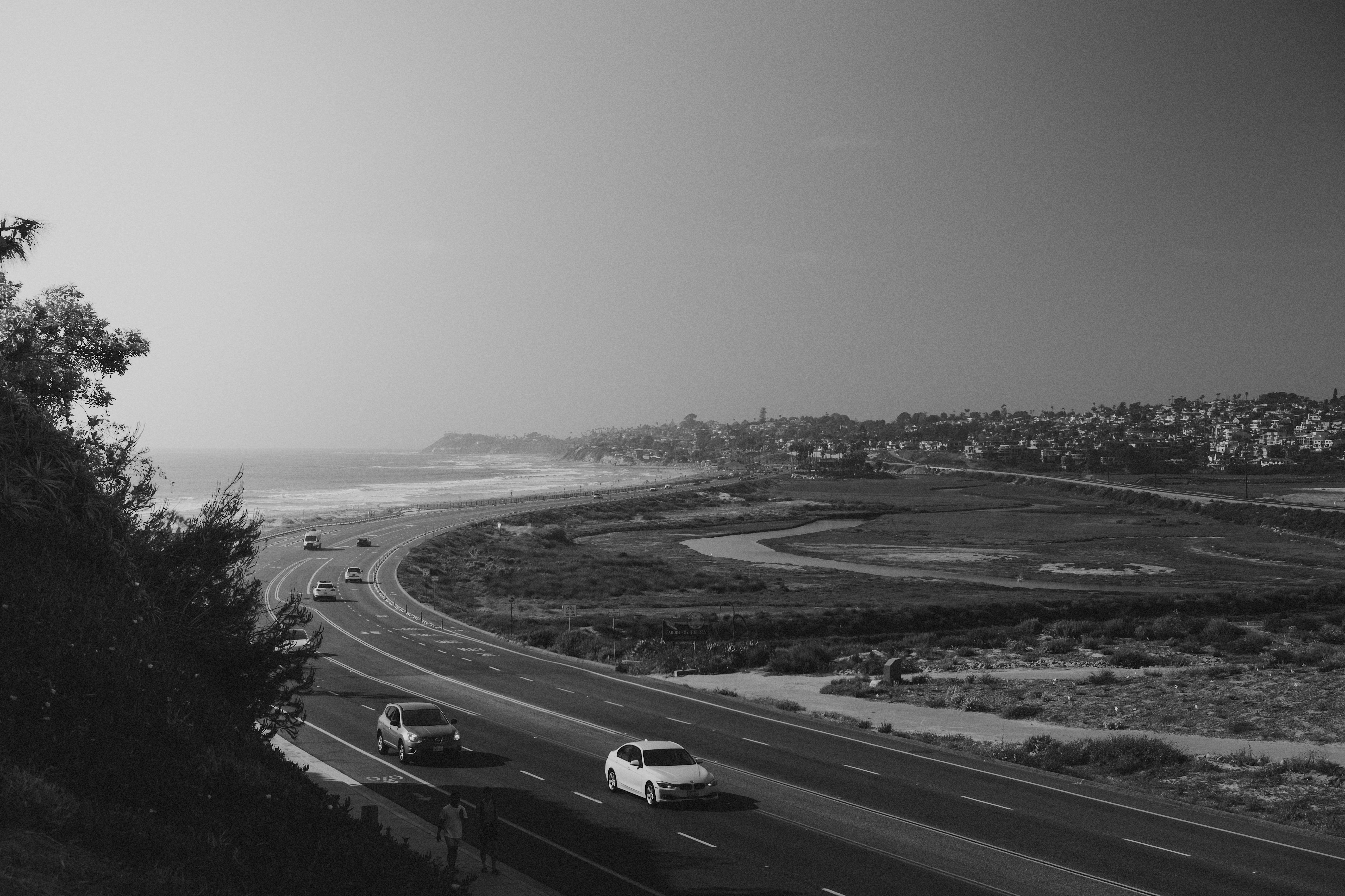 Winding coastal road with vehicles traveling alongside the ocean, framed by lush greenery and distant hills. Black and white tones enhance the tranquil atmosphere.
