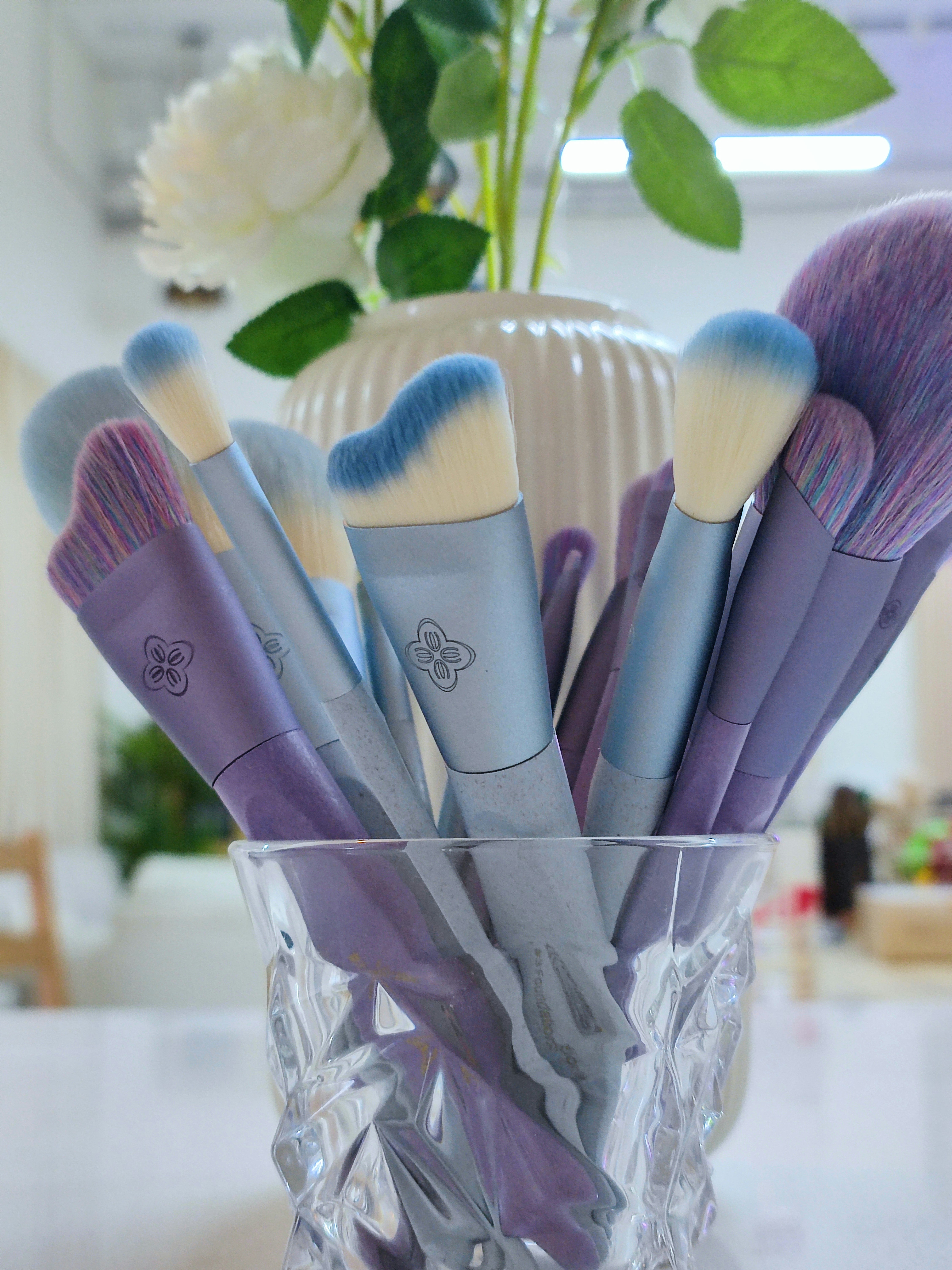A collection of colorful makeup brushes arranged in a crystal vase, surrounded by soft greenery and a hint of floral decor.