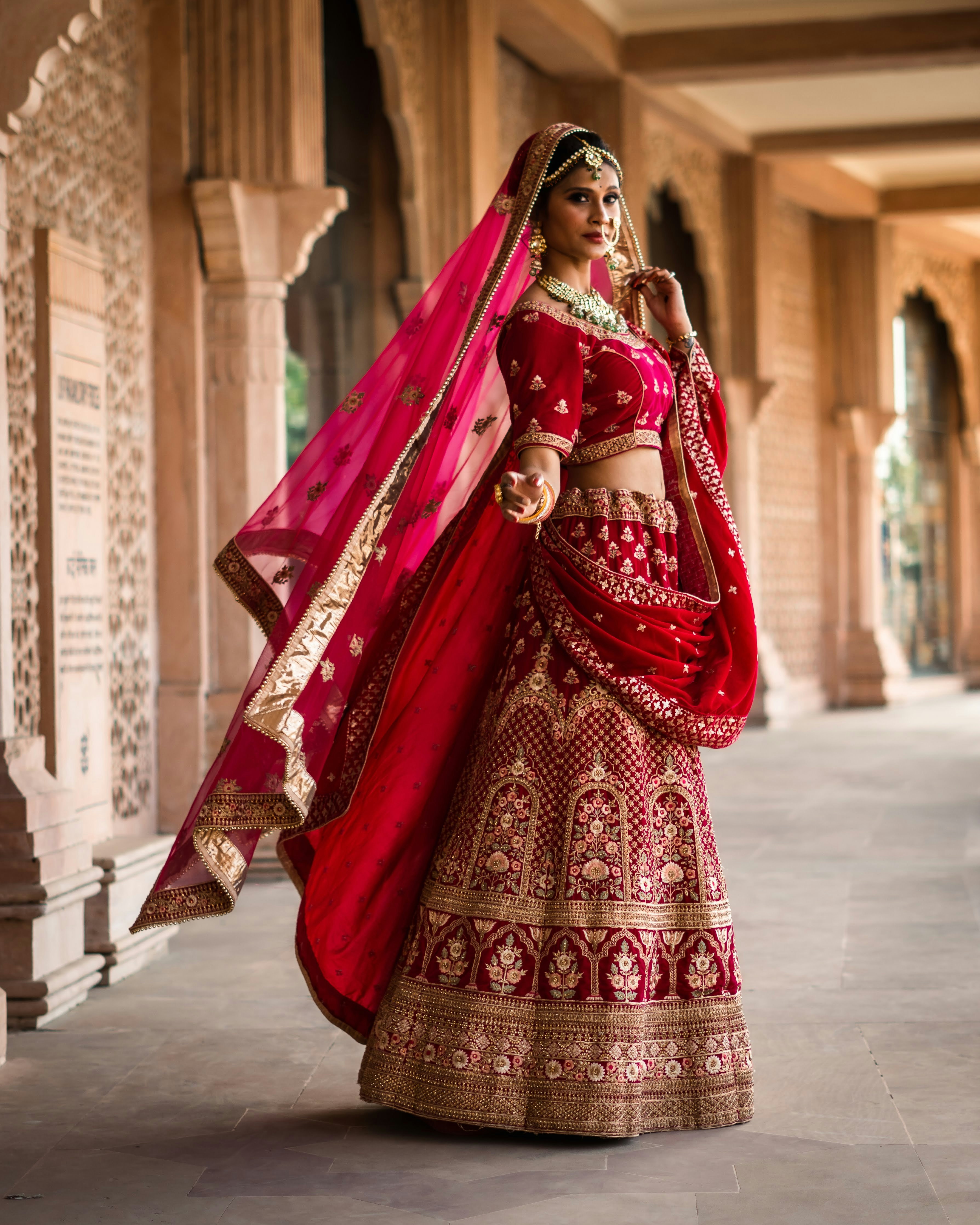 A woman in a vibrant red lehenga and pink dupatta poses gracefully in an ornate architectural setting, showcasing traditional attire. The intricate details of her outfit highlight cultural craftsmanship.
