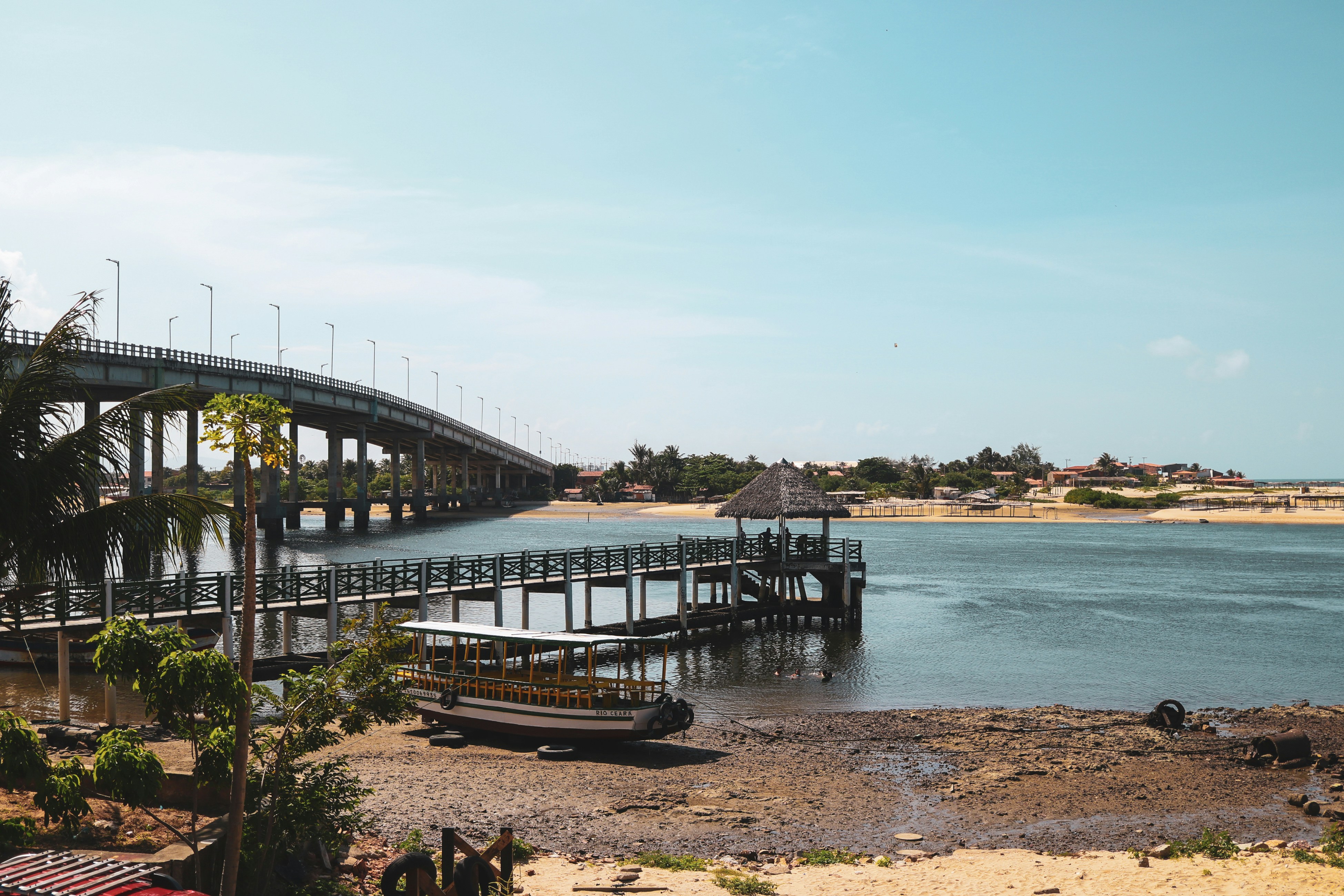 Curved bridge spanning a tranquil waterway beside a sandy beach under a clear sky.