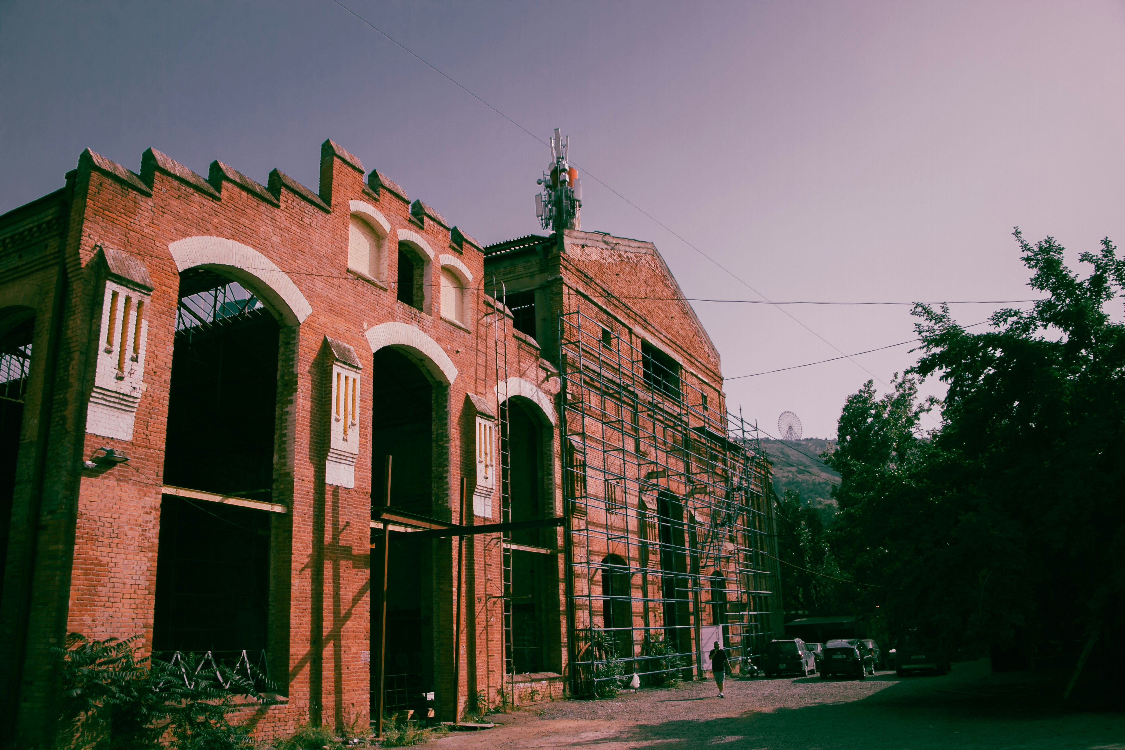Abandoned brick factory with scaffolding and a telecommunications tower amidst a backdrop of trees and a hazy sky.
