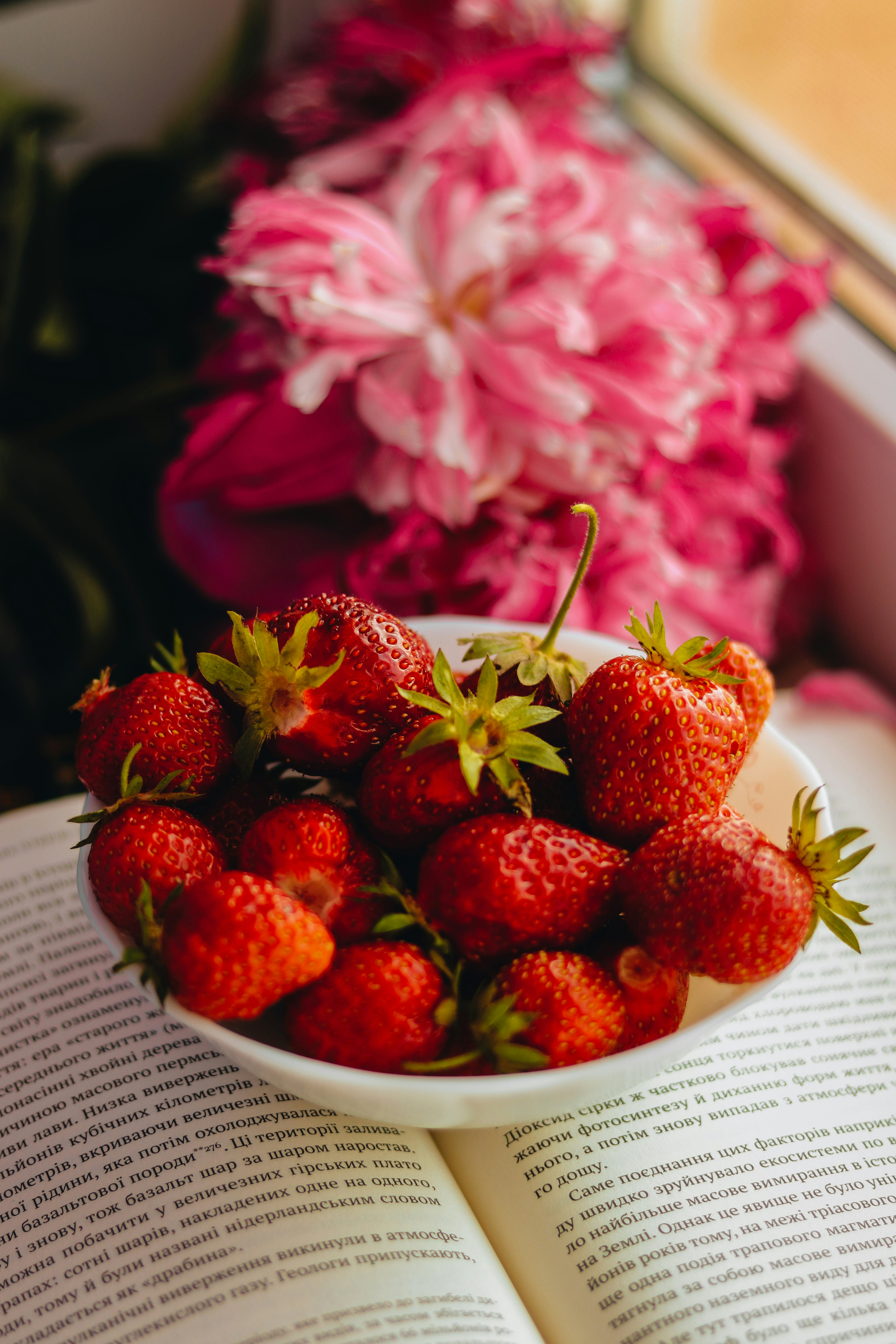 A bowl overflowing with ripe strawberries placed on an open book, with vibrant pink flowers softly blurred in the background.