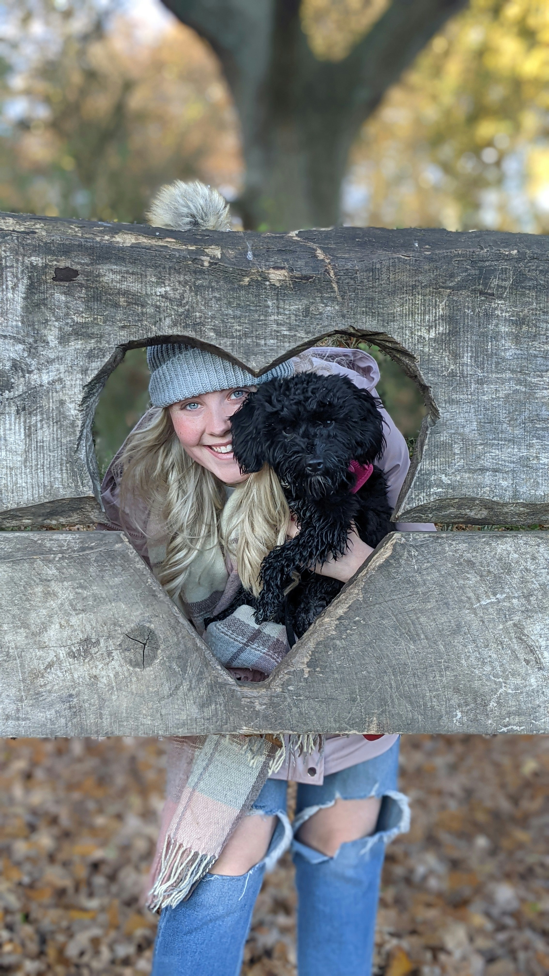 A woman in a gray beanie and scarf holds a fluffy black dog through a heart-shaped cut in a weathered wooden fence, with autumn leaves in the background.
