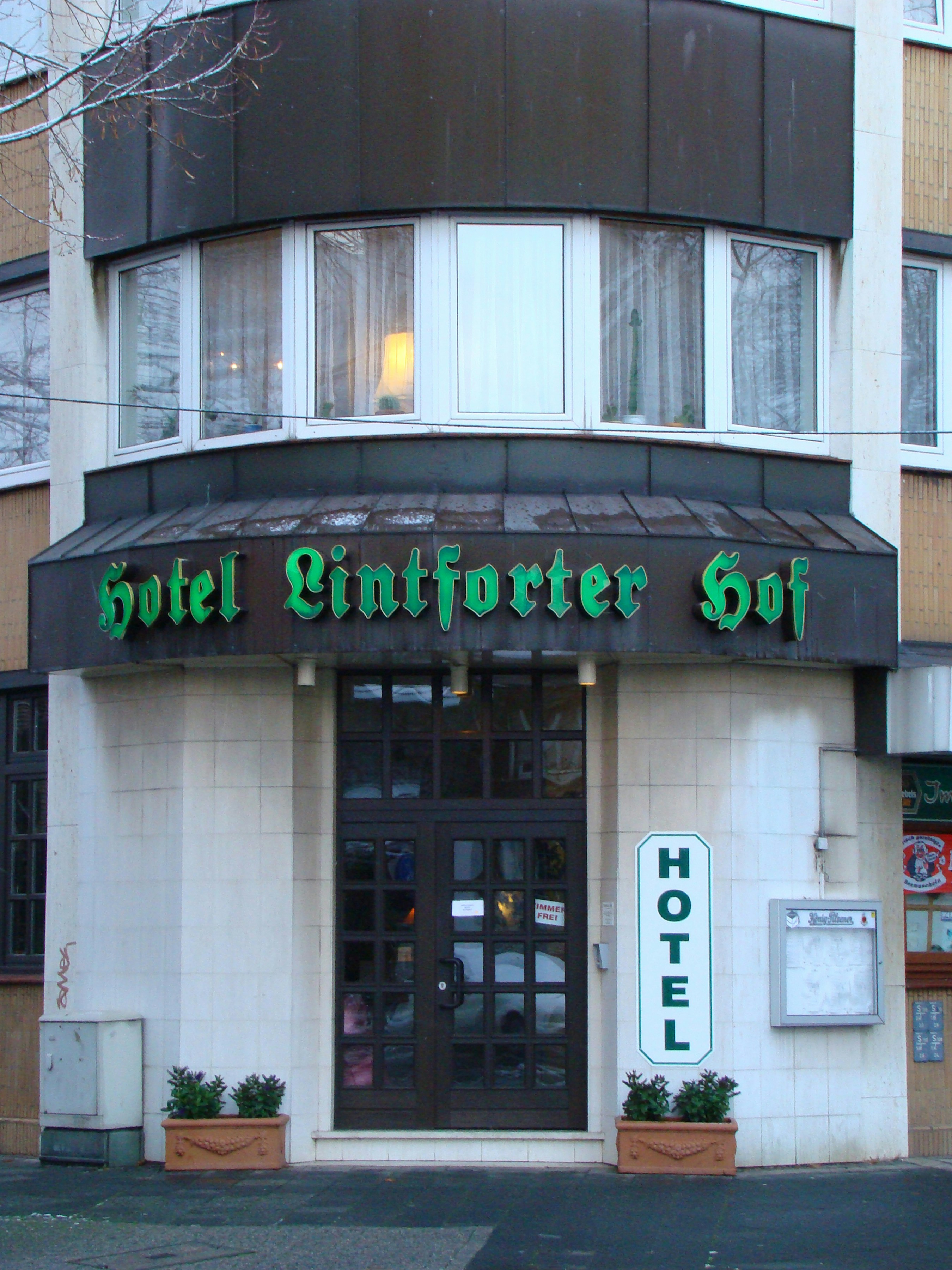 Elegant hotel entrance featuring a distinct curved design and green signage. The inviting atmosphere is accentuated by potted plants at the base.