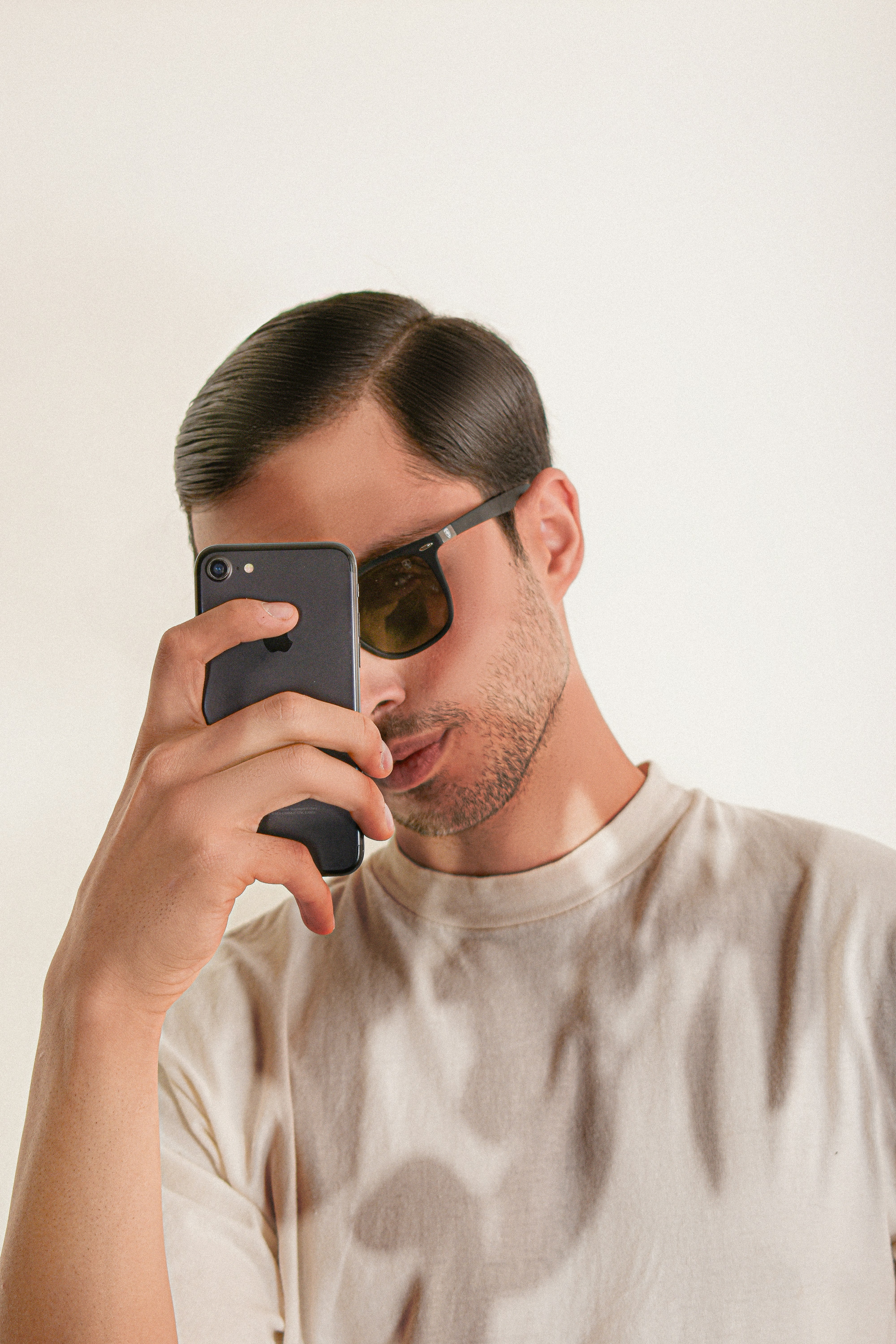 A young man wearing sunglasses and a casual t-shirt holds a smartphone in front of his face, creating an intriguing interplay of light and shadow.