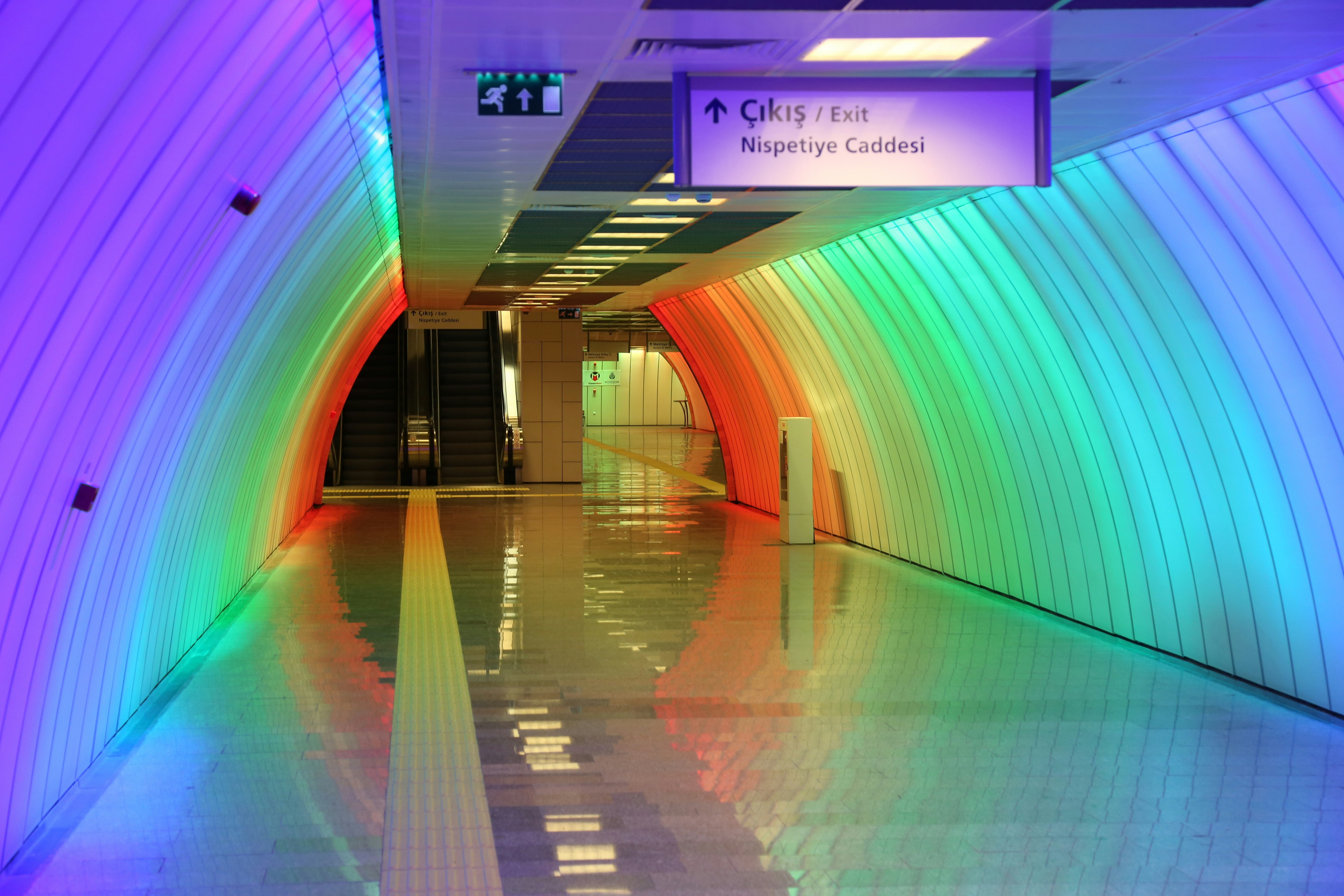 Vibrant illuminated tunnel with a spectrum of colors reflecting on the glossy floor, guiding towards an exit sign.