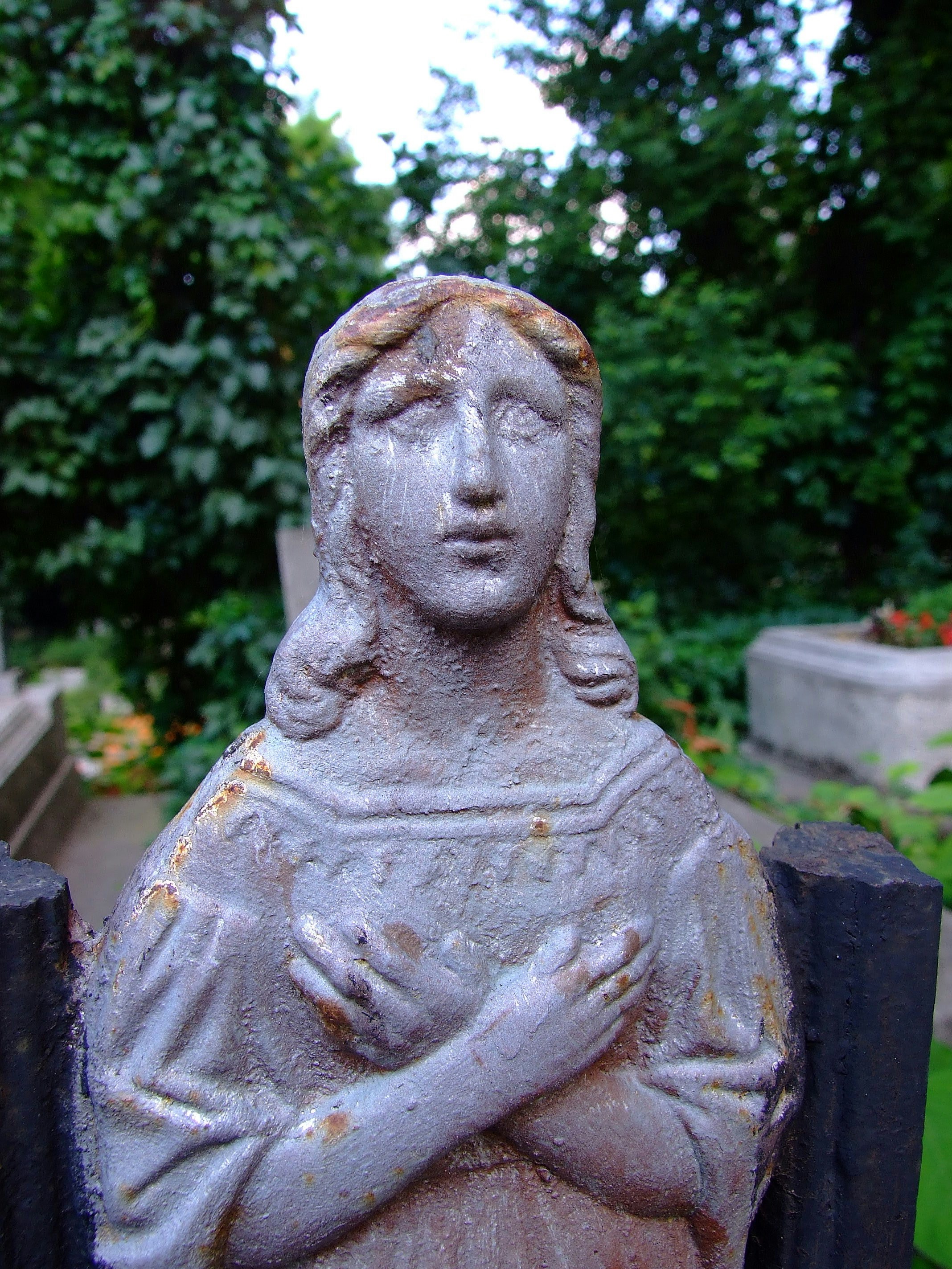 A weathered statue of a figure with clasped hands, set against a backdrop of greenery in a serene cemetery.