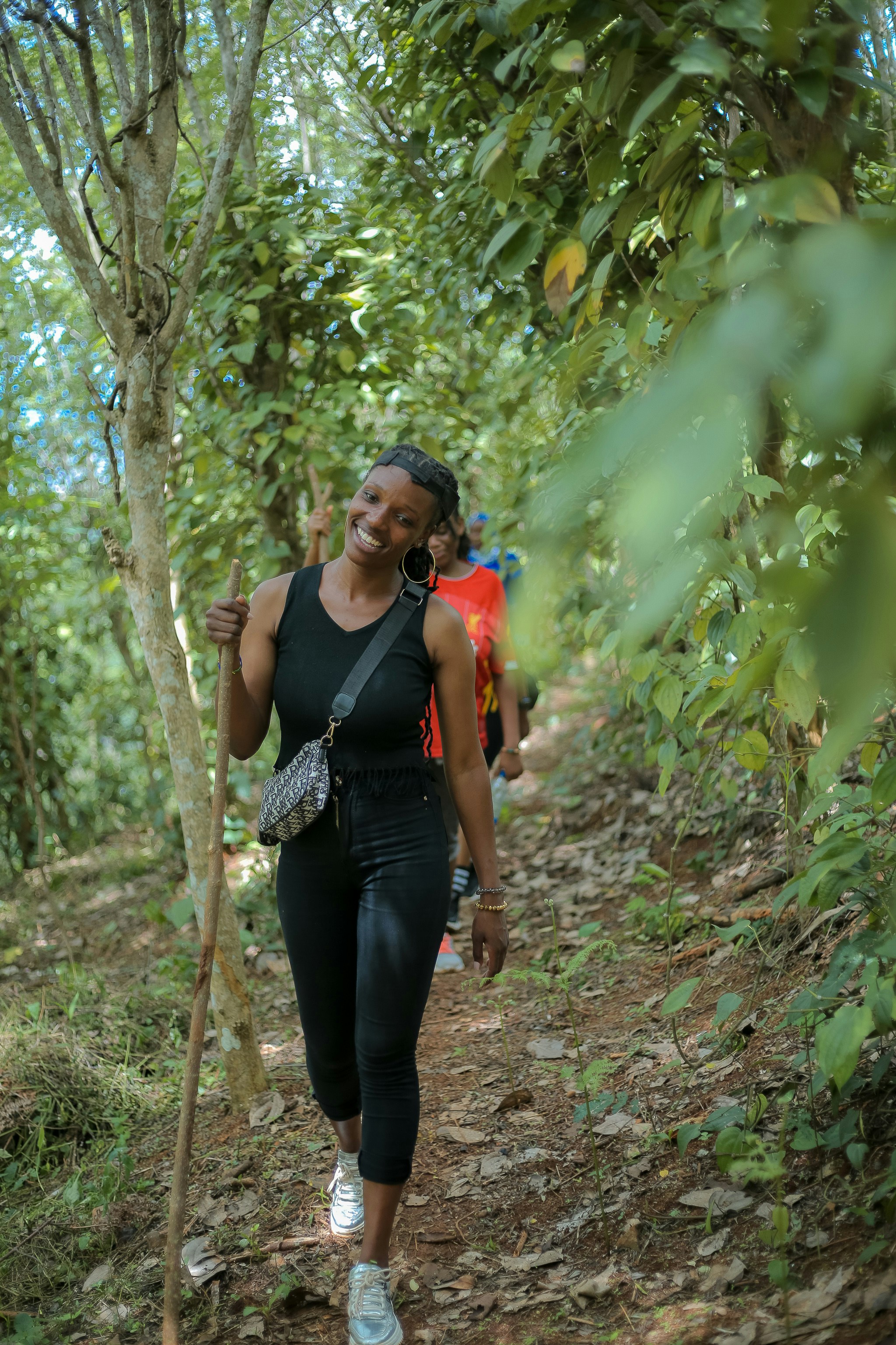 Woman hiking along a lush forest path, holding a walking stick and smiling under dappled sunlight.