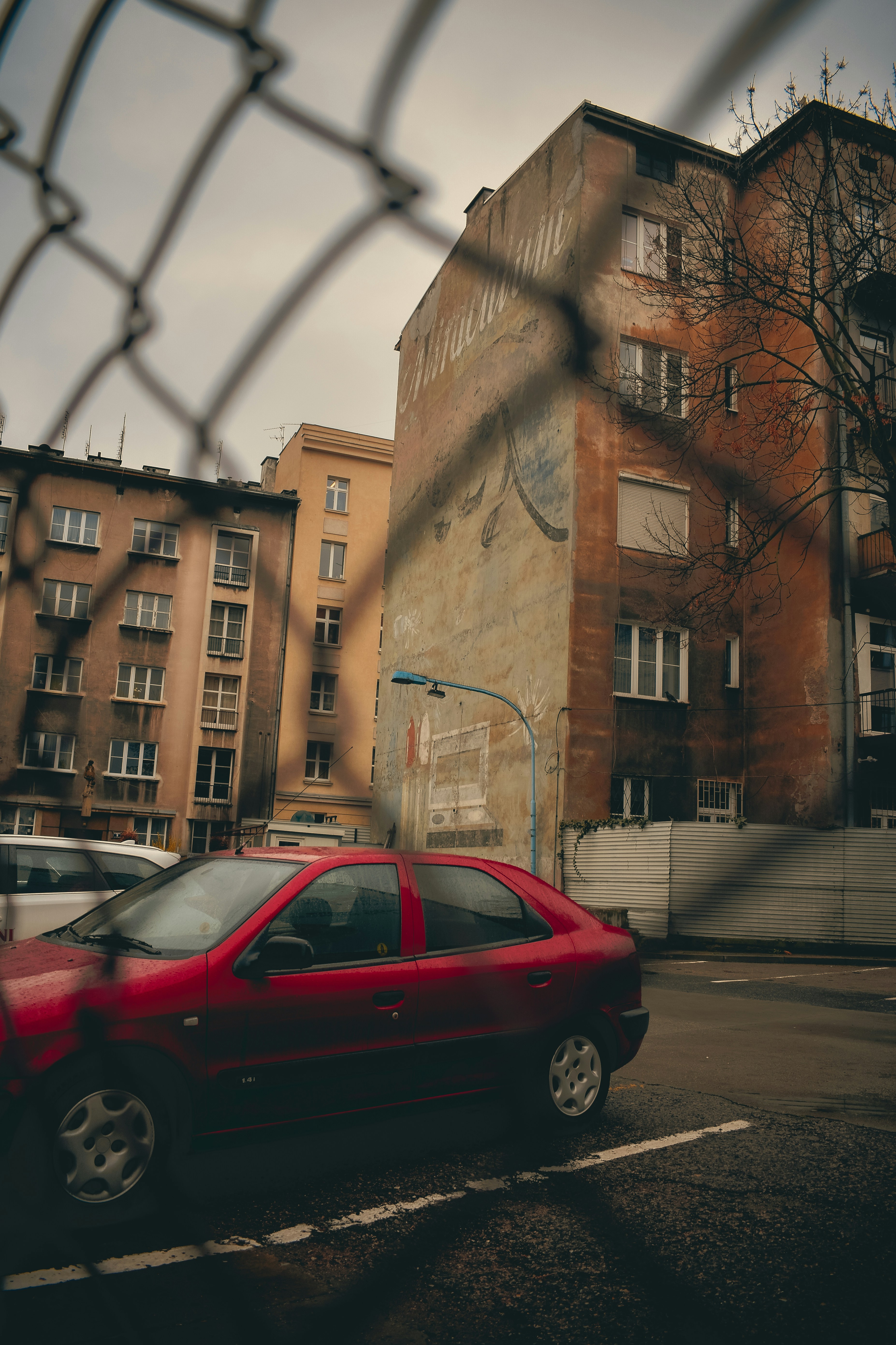 Red sedan parked in a dim city lot with a mural-adorned, weathered building in the background; a chain-link fence foreground adds a gritty urban mood.