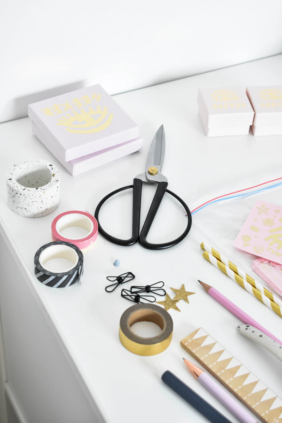 Overhead view of a bright white desk with scissors, washi tape, paper clips, pencils, and small decorative boxes arranged in a tidy craft flat lay.