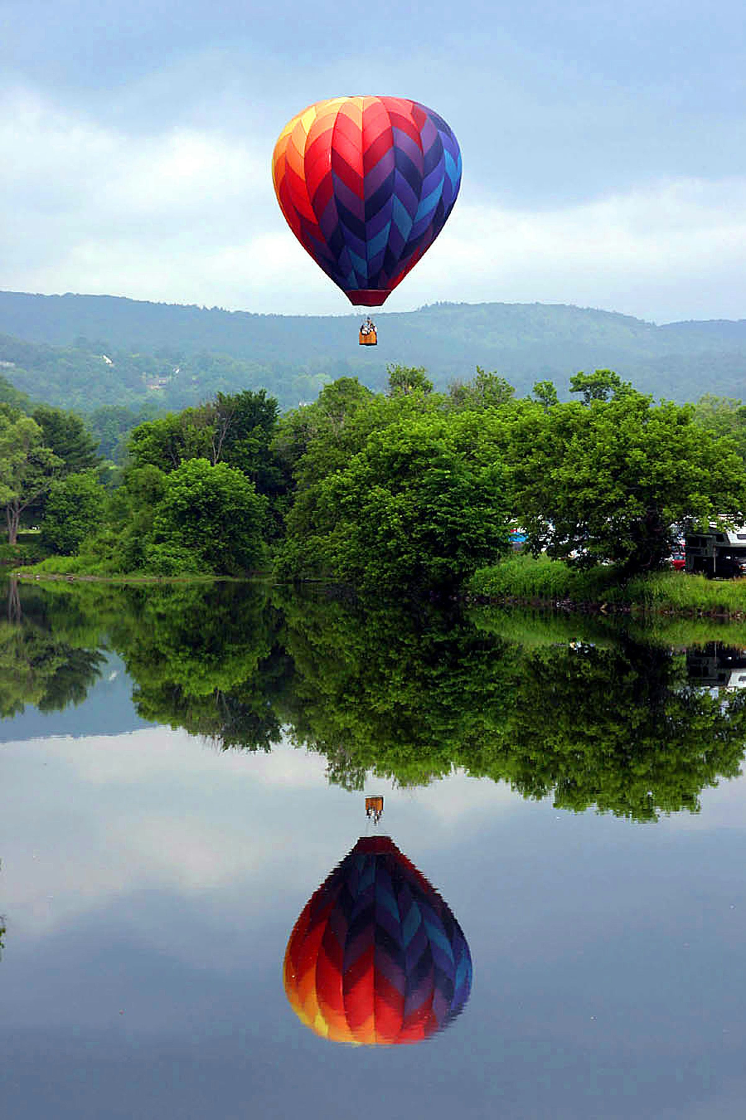 Colorful chevron-patterned hot air balloon drifts above a tree-lined lake, its reflection perfectly mirrored on the water.