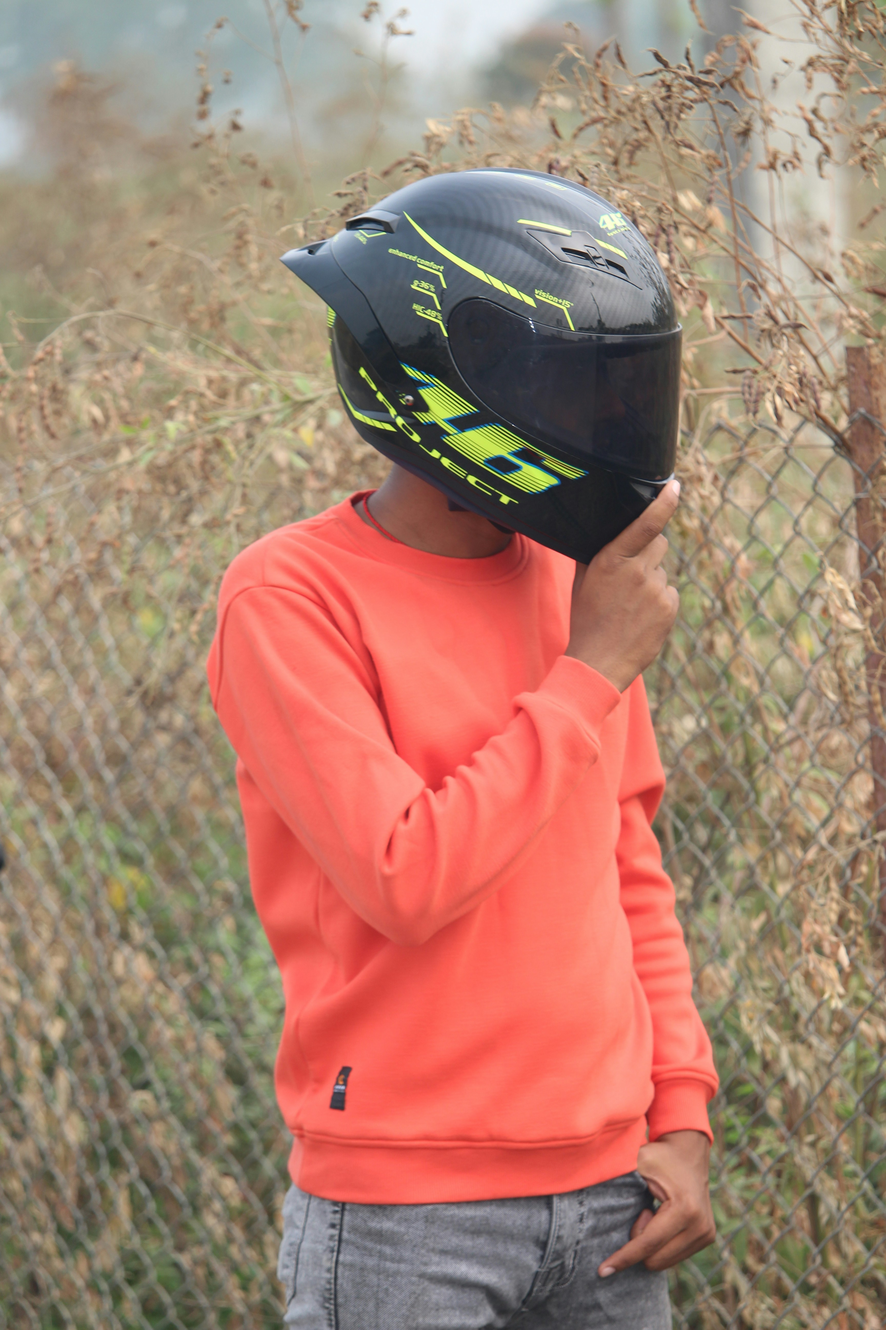 A person in an orange sweatshirt holds a black motorcycle helmet, partially obscuring their face, set against a backdrop of dried foliage and a chain-link fence.