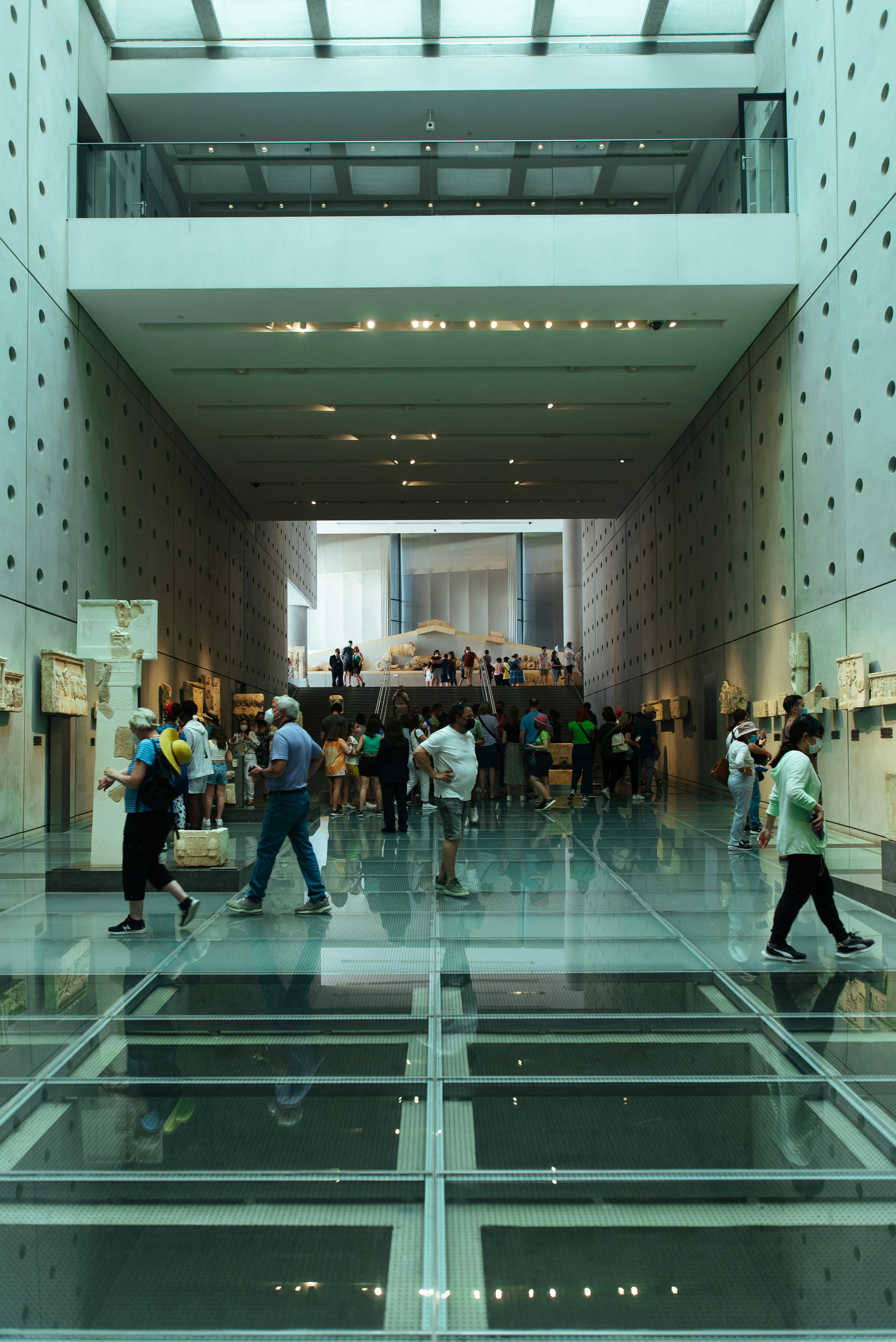 Visitors explore a modern museum space filled with ancient artifacts and contemporary architecture. The glass floor adds a unique perspective to the exhibits.