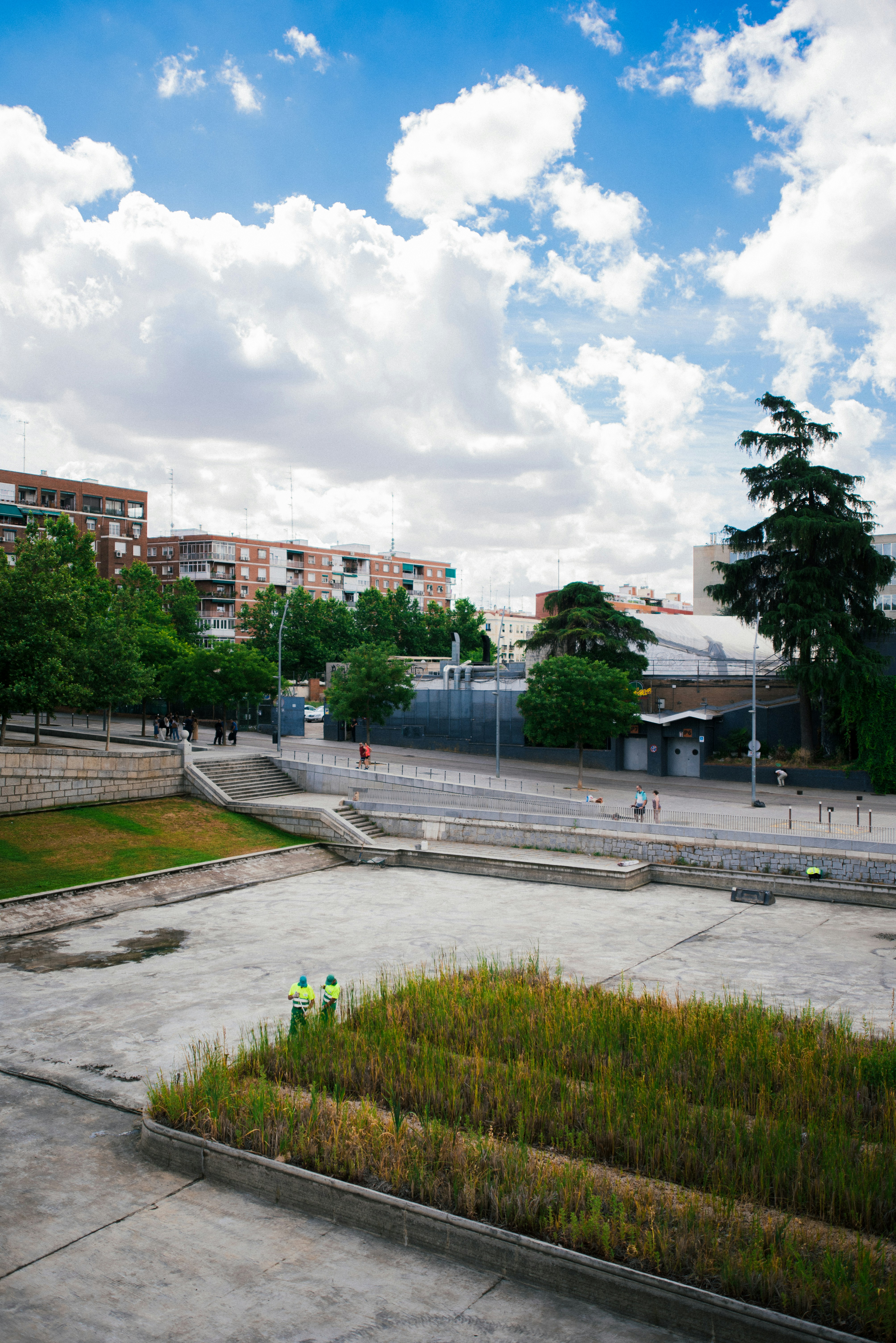 Two workers tending to a patch of wild grass in an urban park setting, surrounded by modern architecture and vibrant greenery.