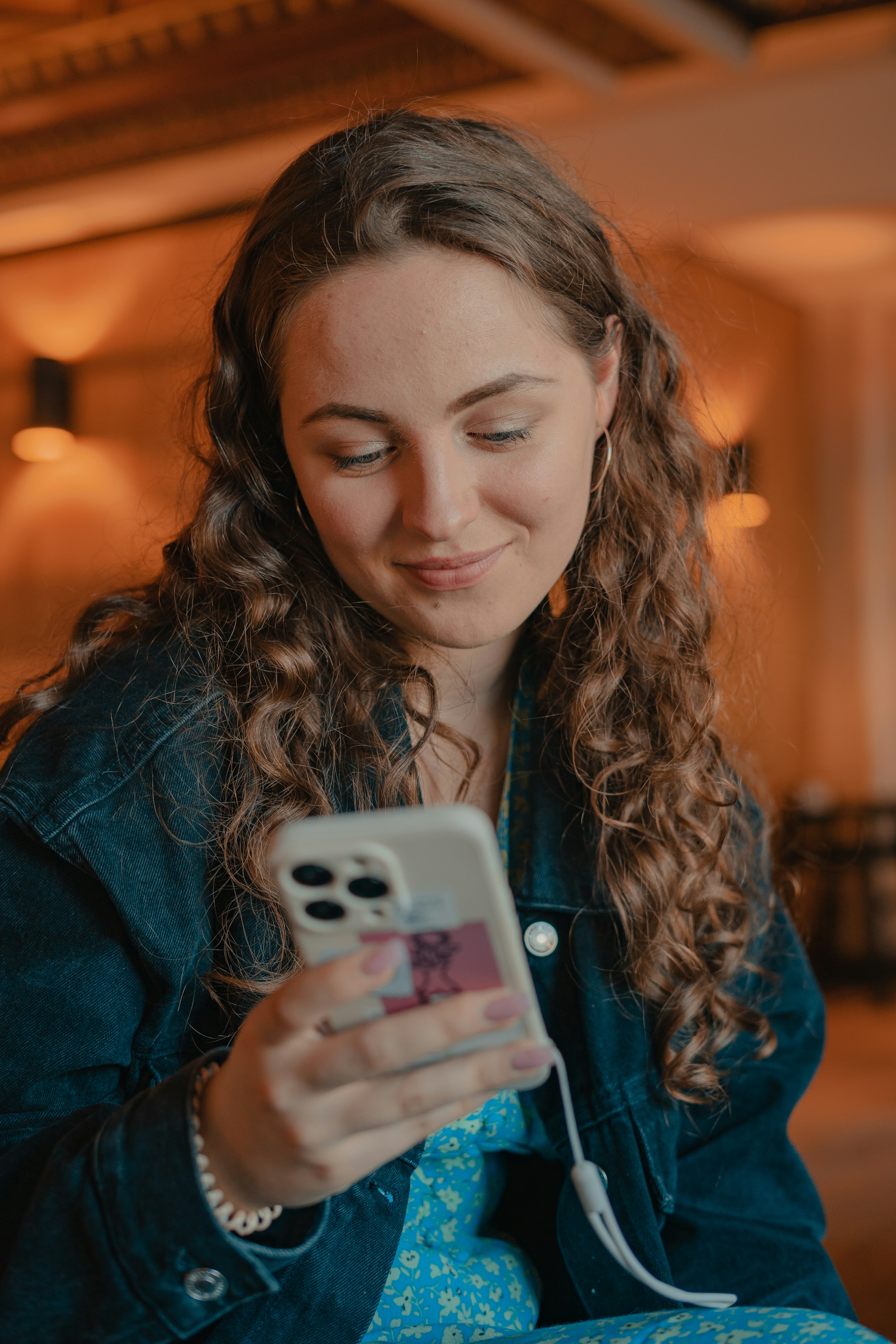 Young woman with curly hair engrossed in her smartphone, wearing a denim jacket in a softly lit café setting.