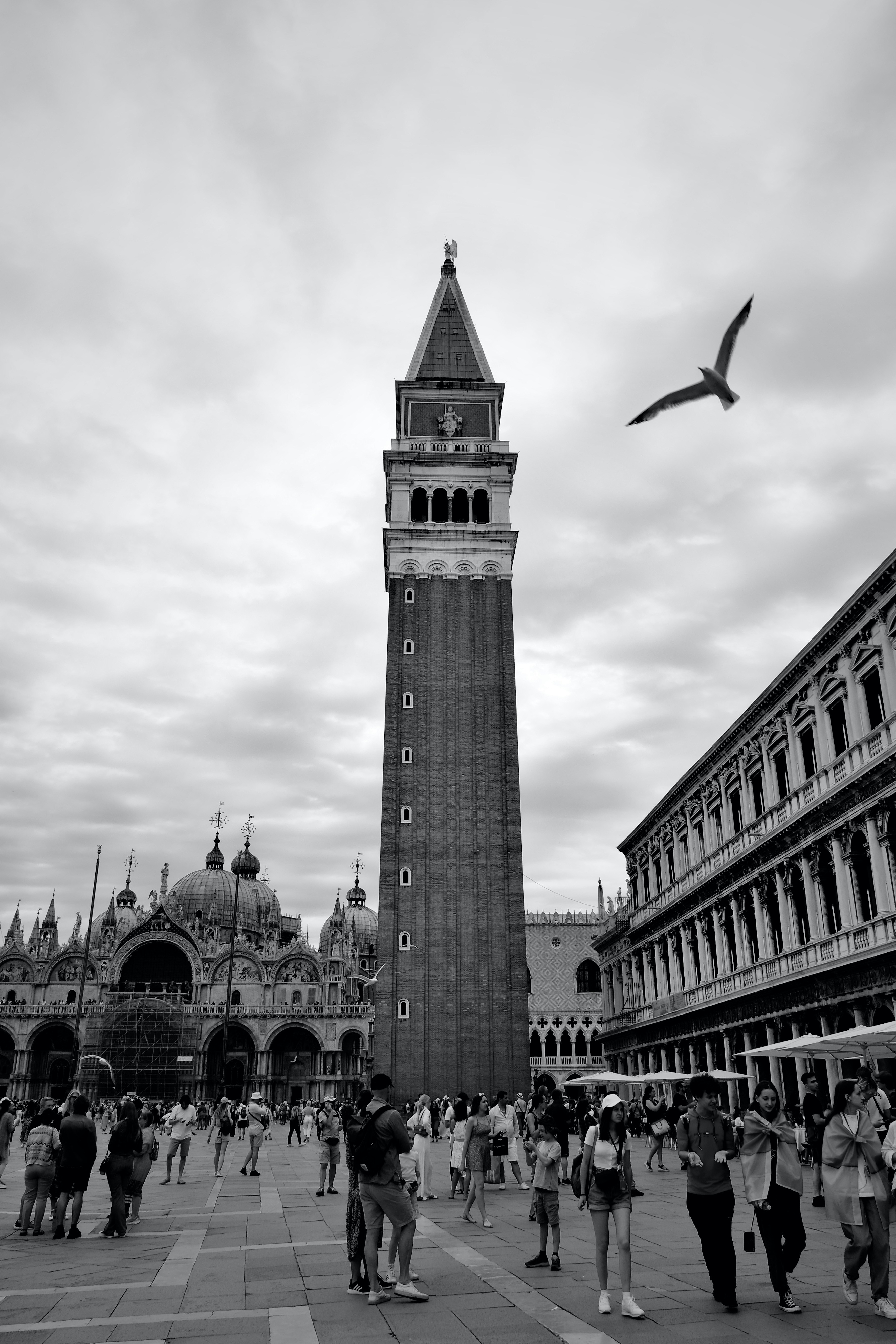 The iconic Campanile of St. Mark's Basilica stands tall amidst a bustling square, with visitors exploring the historic site under a moody sky.