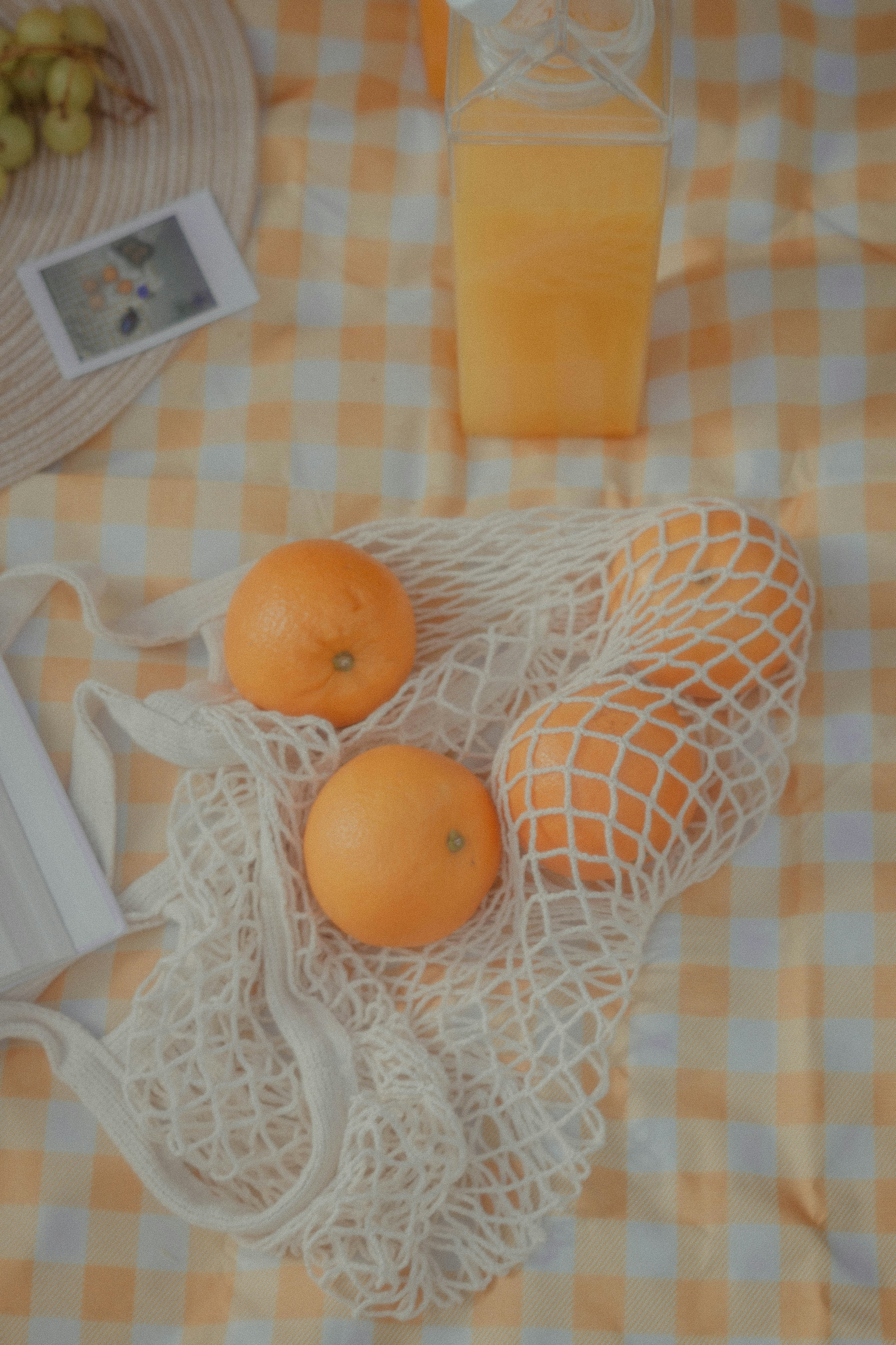 Fresh oranges nestled in a netted bag on a checkered tablecloth, accompanied by a glass of orange juice and a vintage photo. 
