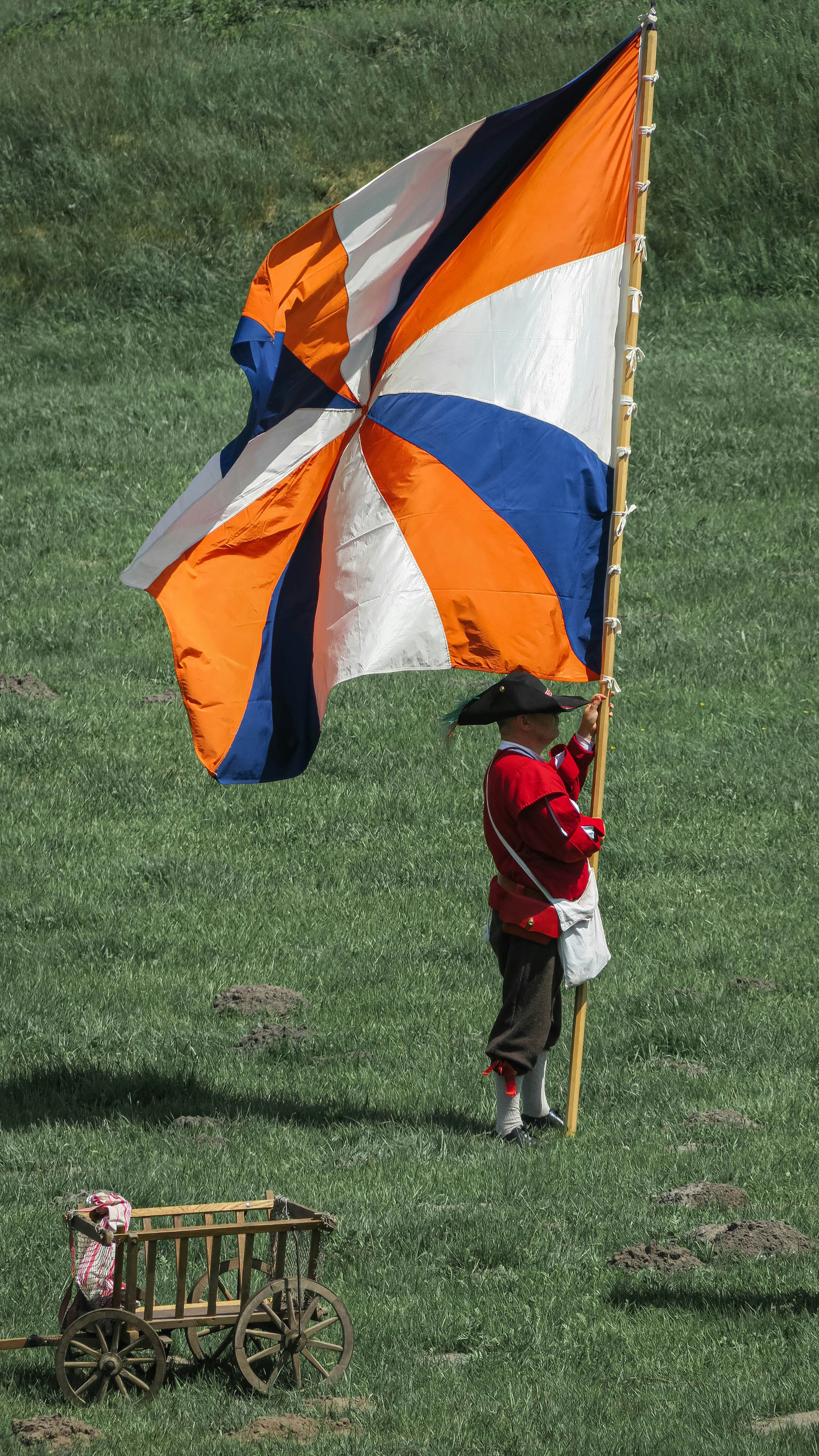 A historical reenactor stands proudly, holding a vibrant flag with orange, blue, and white sections, while a wooden cart rests nearby on the grassy field.
