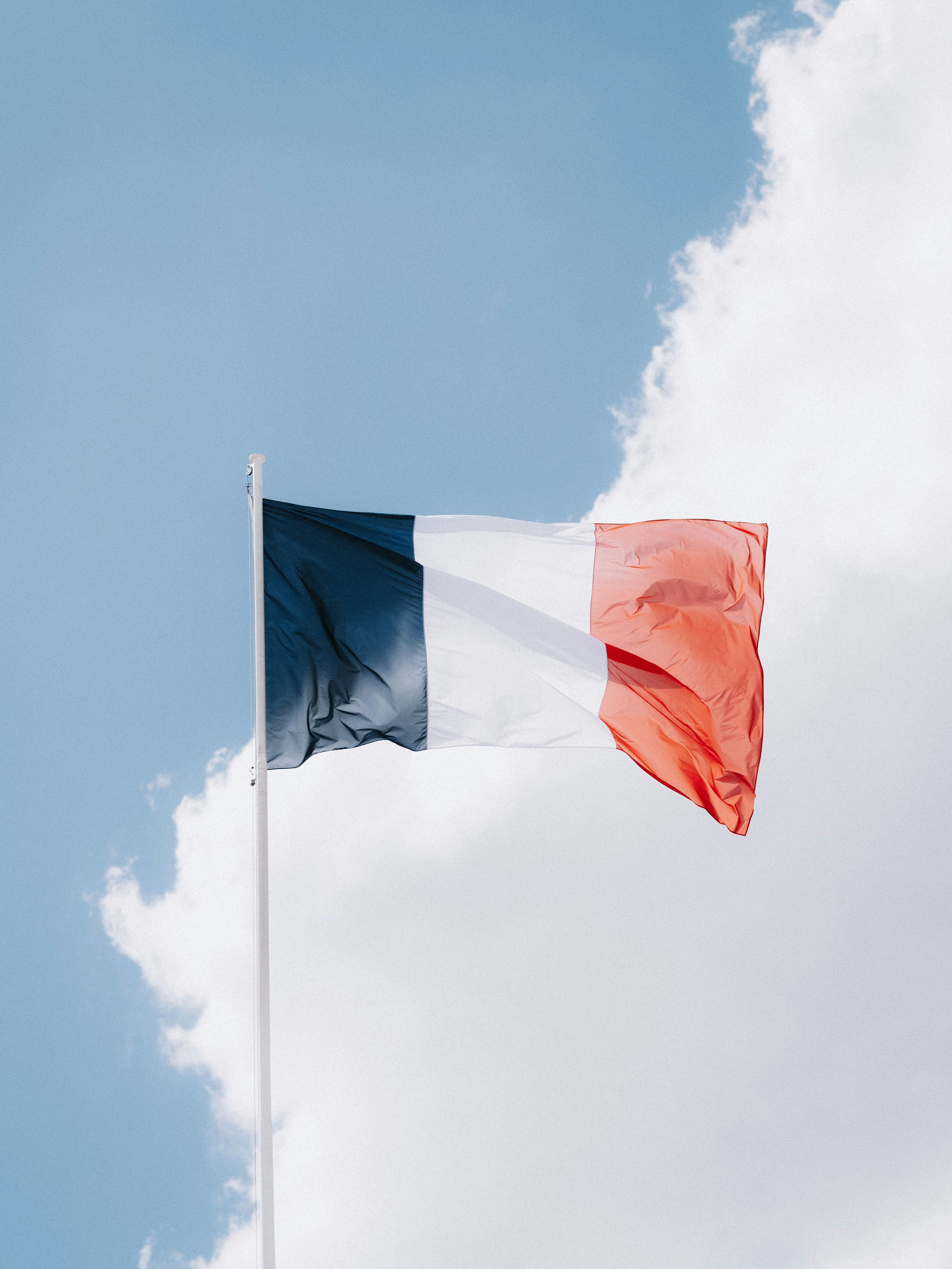 French flag billowing against a bright blue sky, showcasing its iconic tricolor design. The clouds add depth to the scene.
