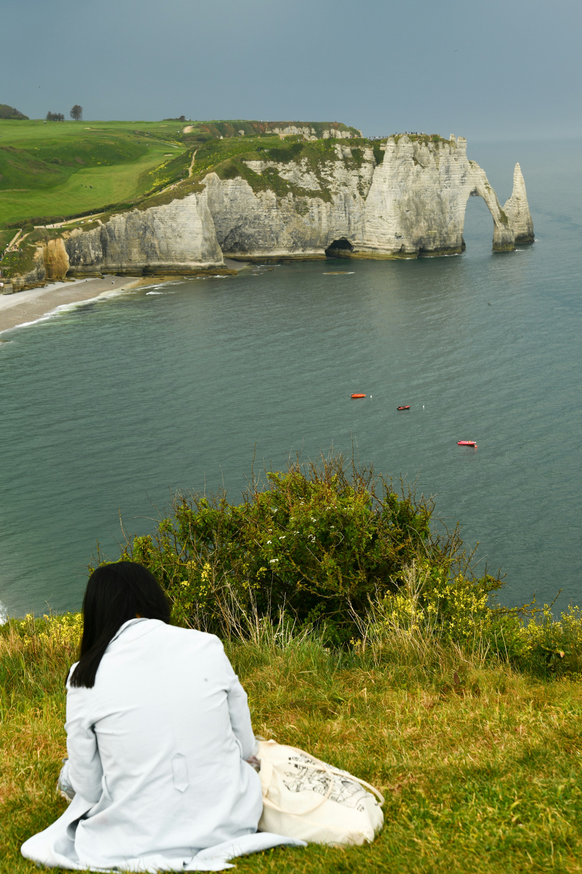 A person in a white coat sits on a grassy cliff overlooking turquoise water, with white chalk cliffs forming a sea arch in the distance.