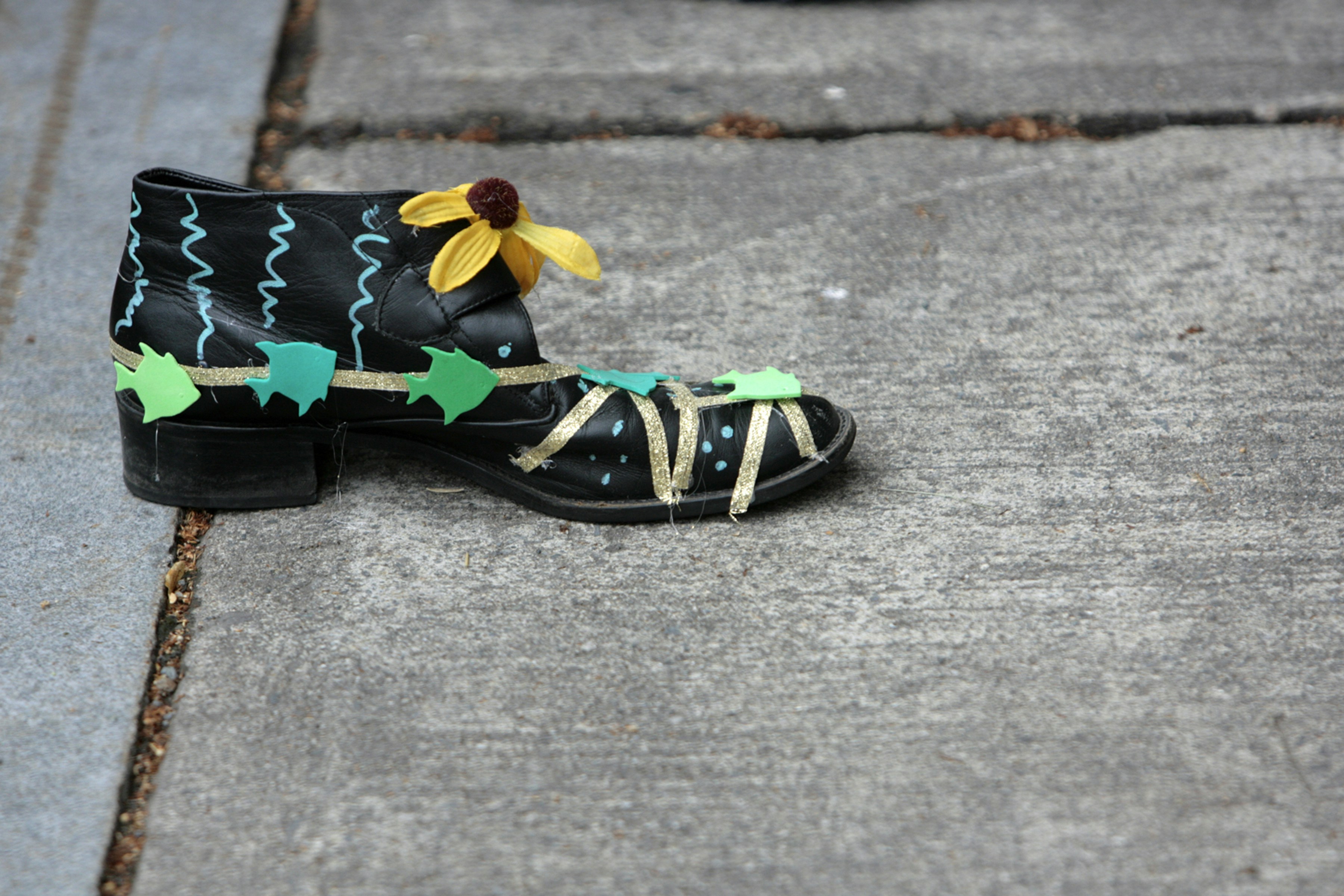 A creatively decorated black shoe adorned with a yellow flower and green fish cutouts, resting on a concrete surface.