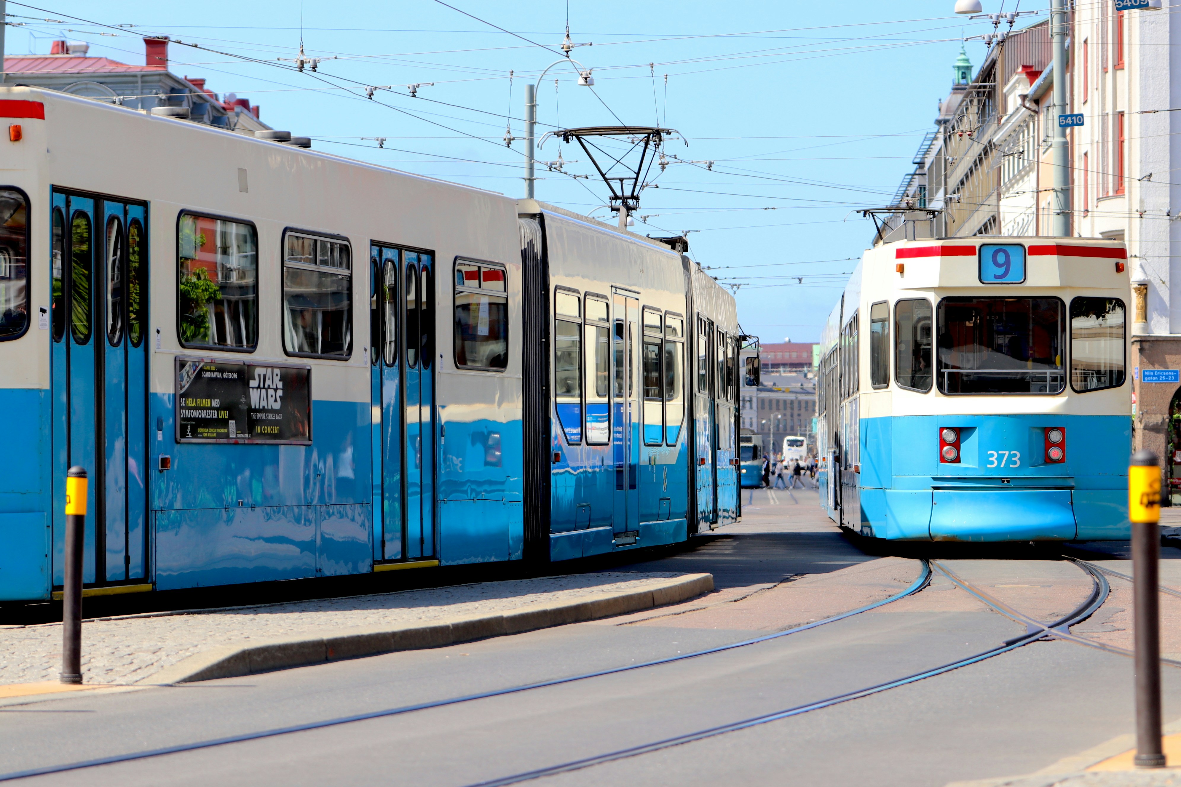 Two trams navigate a bustling city intersection, showcasing vibrant blue colors against a clear sky.