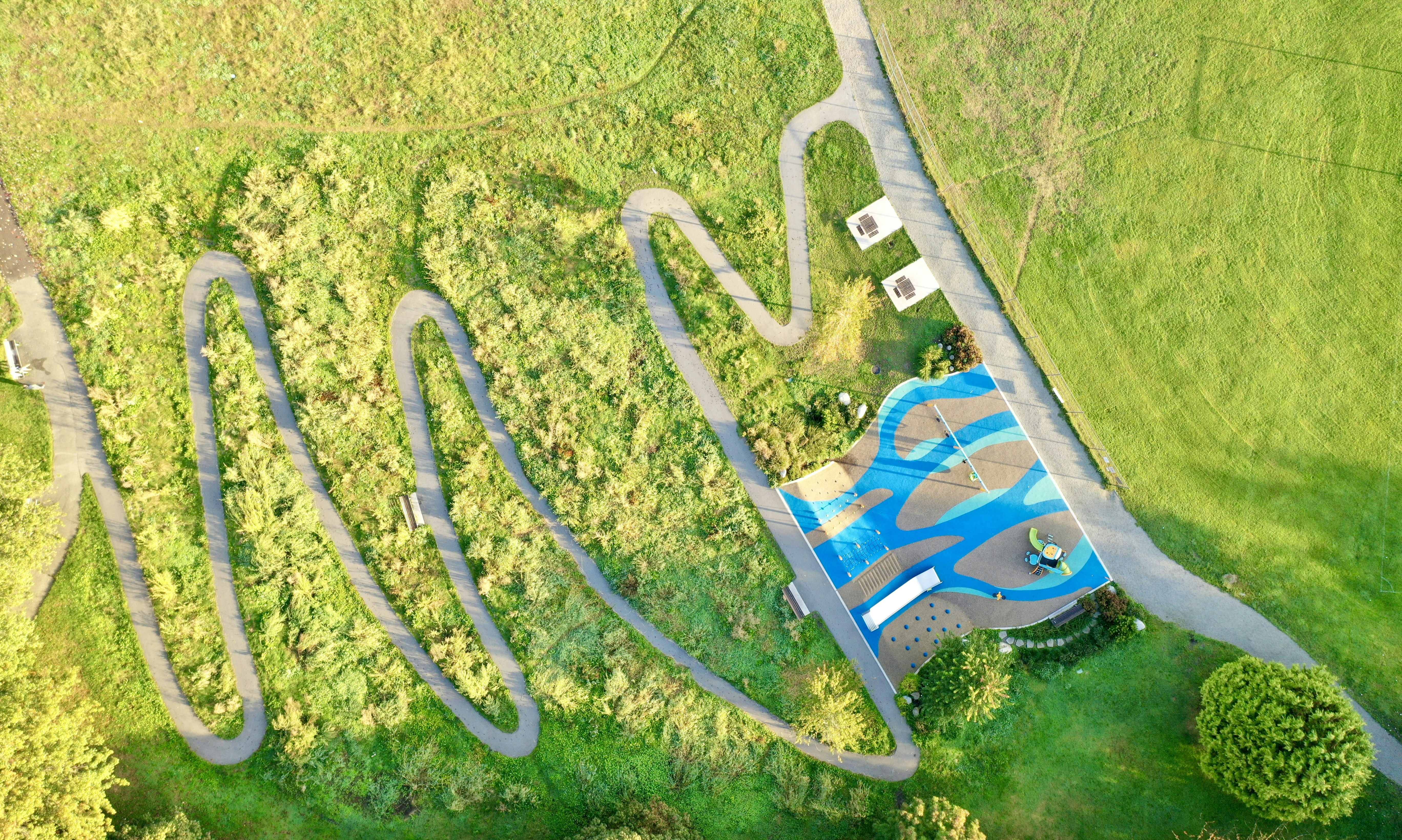 Aerial view of a playground featuring a winding path and vibrant blue play area surrounded by greenery. Benches are positioned nearby for relaxation.