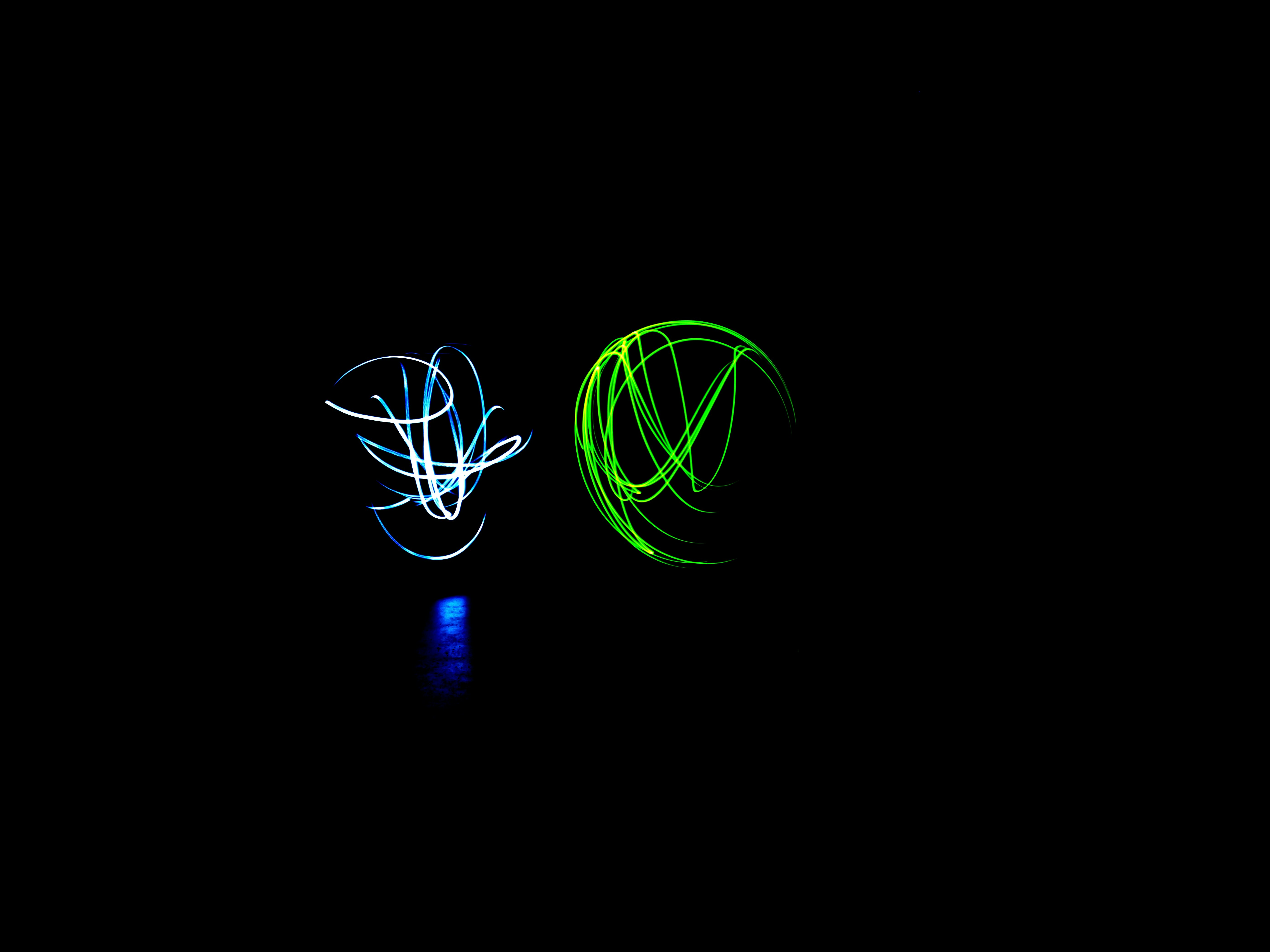 Two neon light trails—blue-white on the left and vivid green on the right—trace abstract spirals against a black backdrop. A minimalist long-exposure photograph emphasizing motion and color.