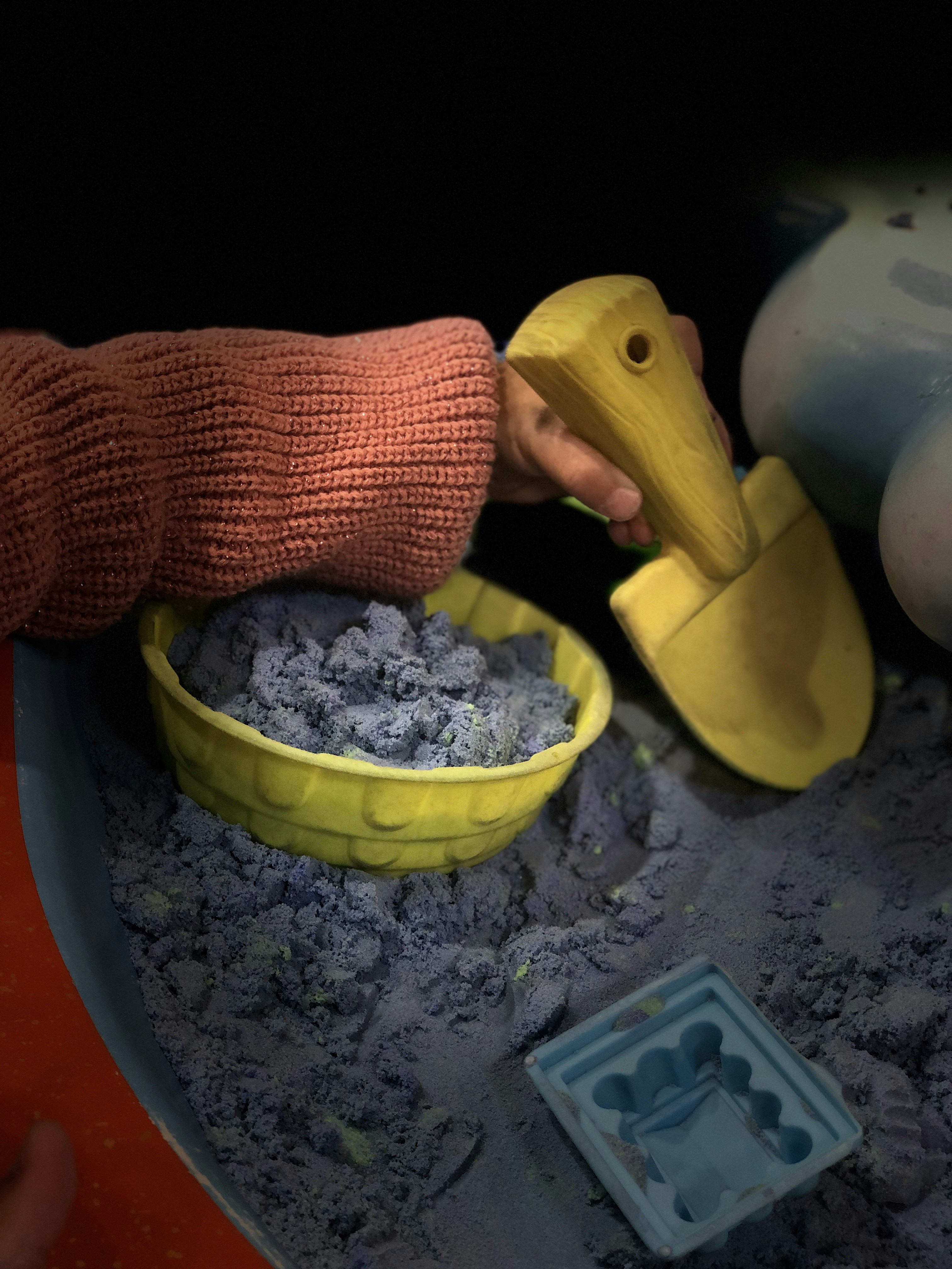 Grip and Glow: A Hand Guides a Yellow Sand Shovel Over Blue Sand in a Dim Sandbox