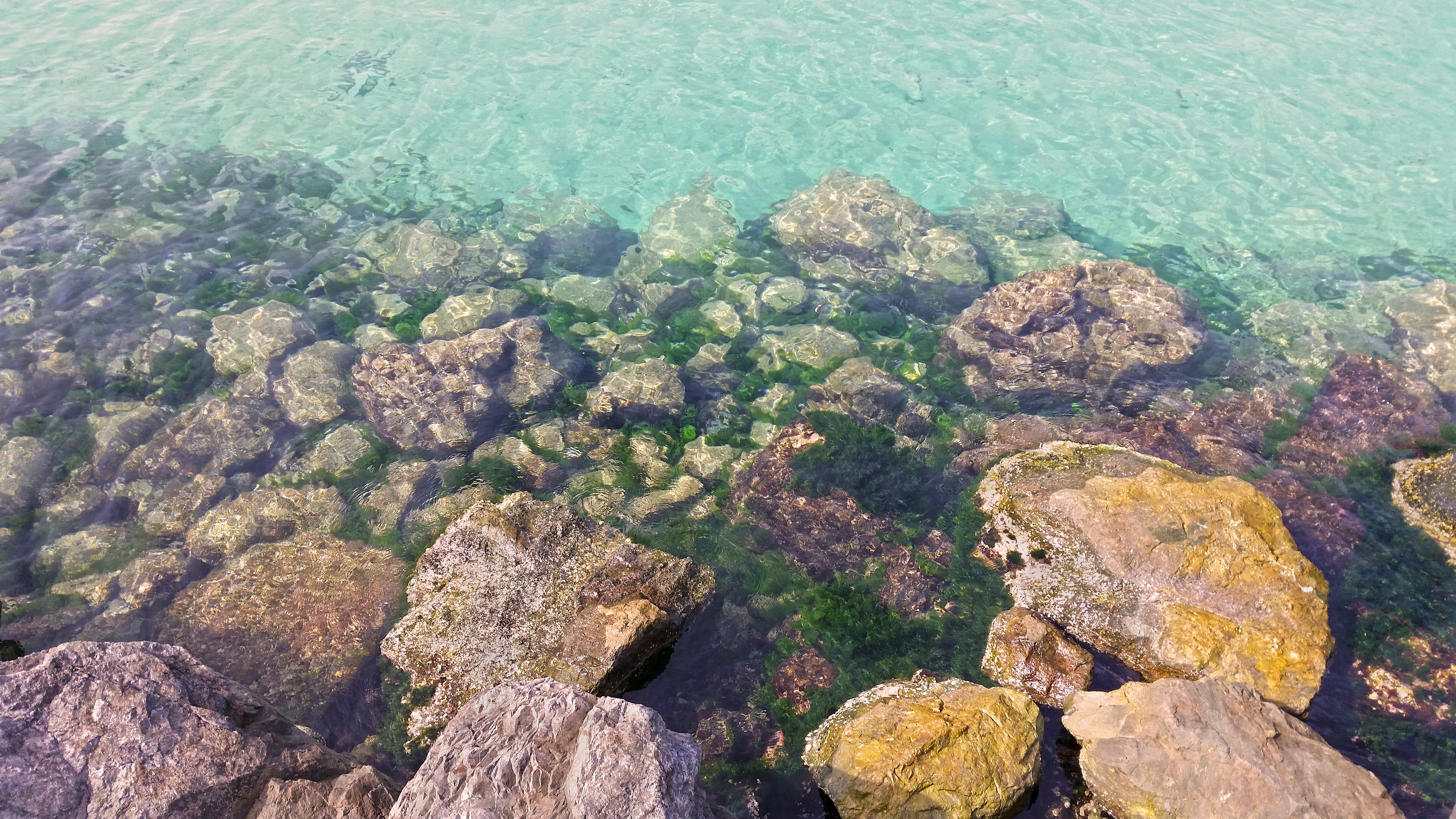 Cluster of sunlit rocks protrudes from clear turquoise water along a rocky shoreline.