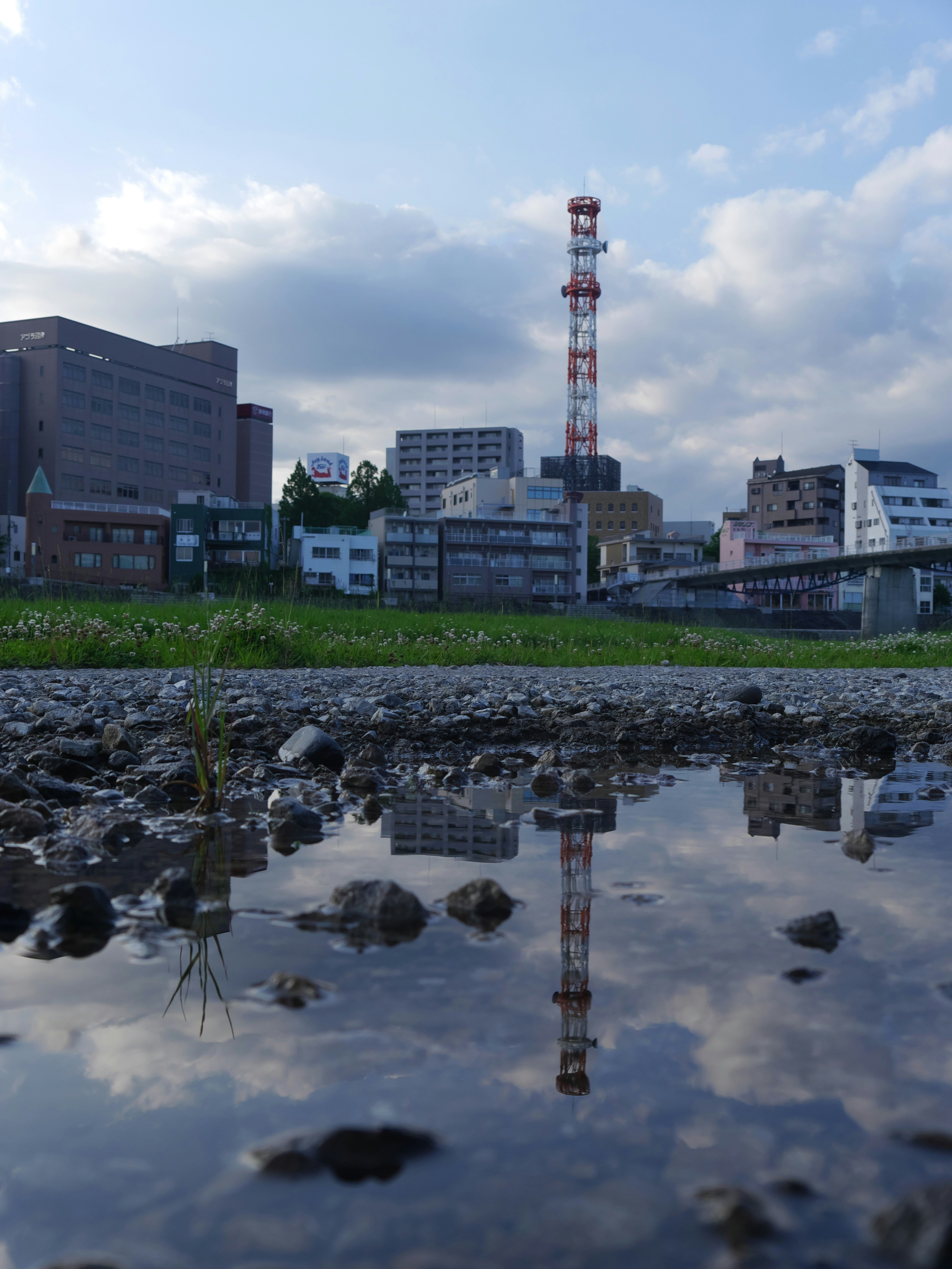 Reflection of a communication tower and city buildings in a puddle, framed by gravel and grass. The scene captures a blend of urban architecture and natural elements.