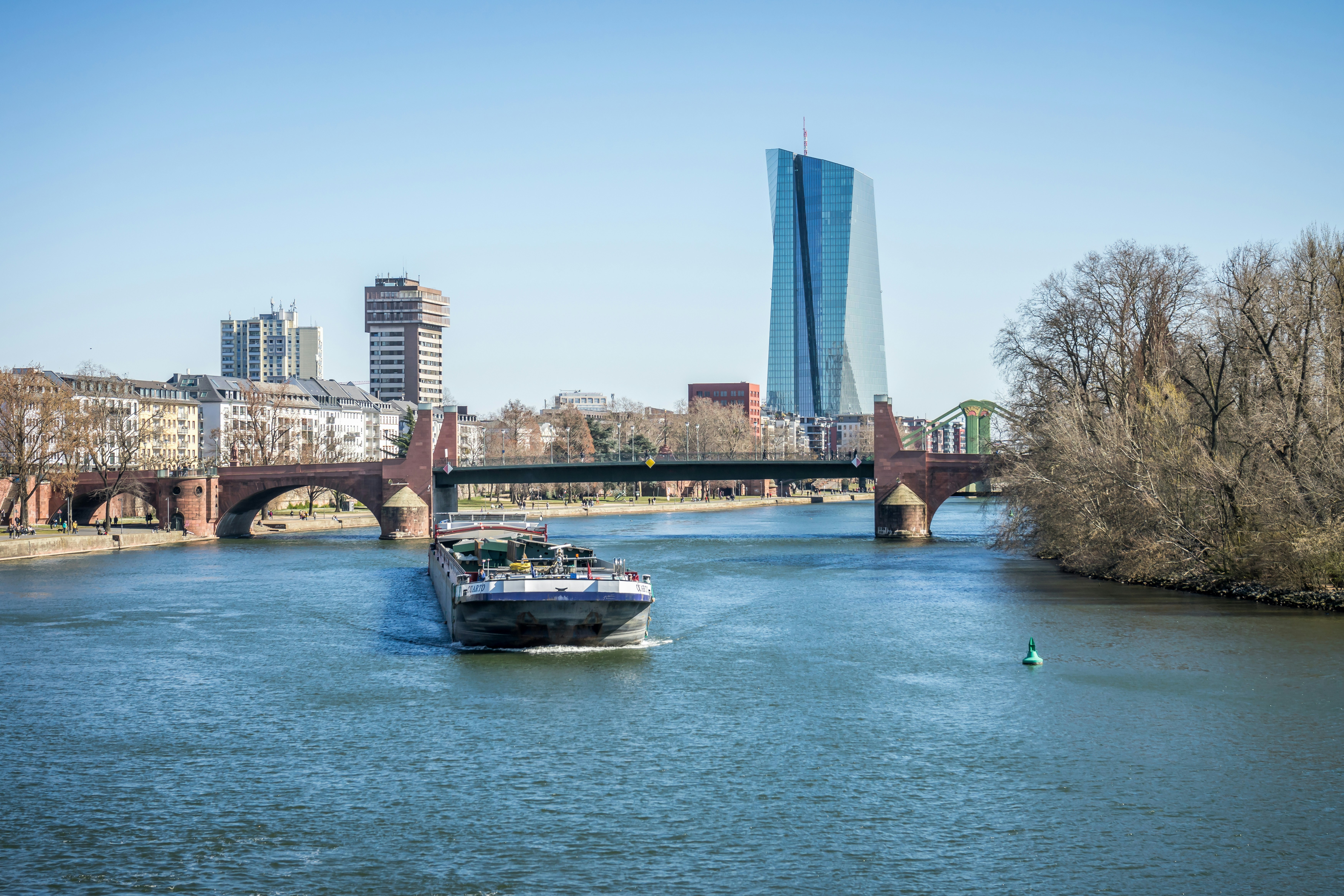 A passenger boat glides along a broad river past a brick arch bridge, with a tall blue glass tower rising in the background.