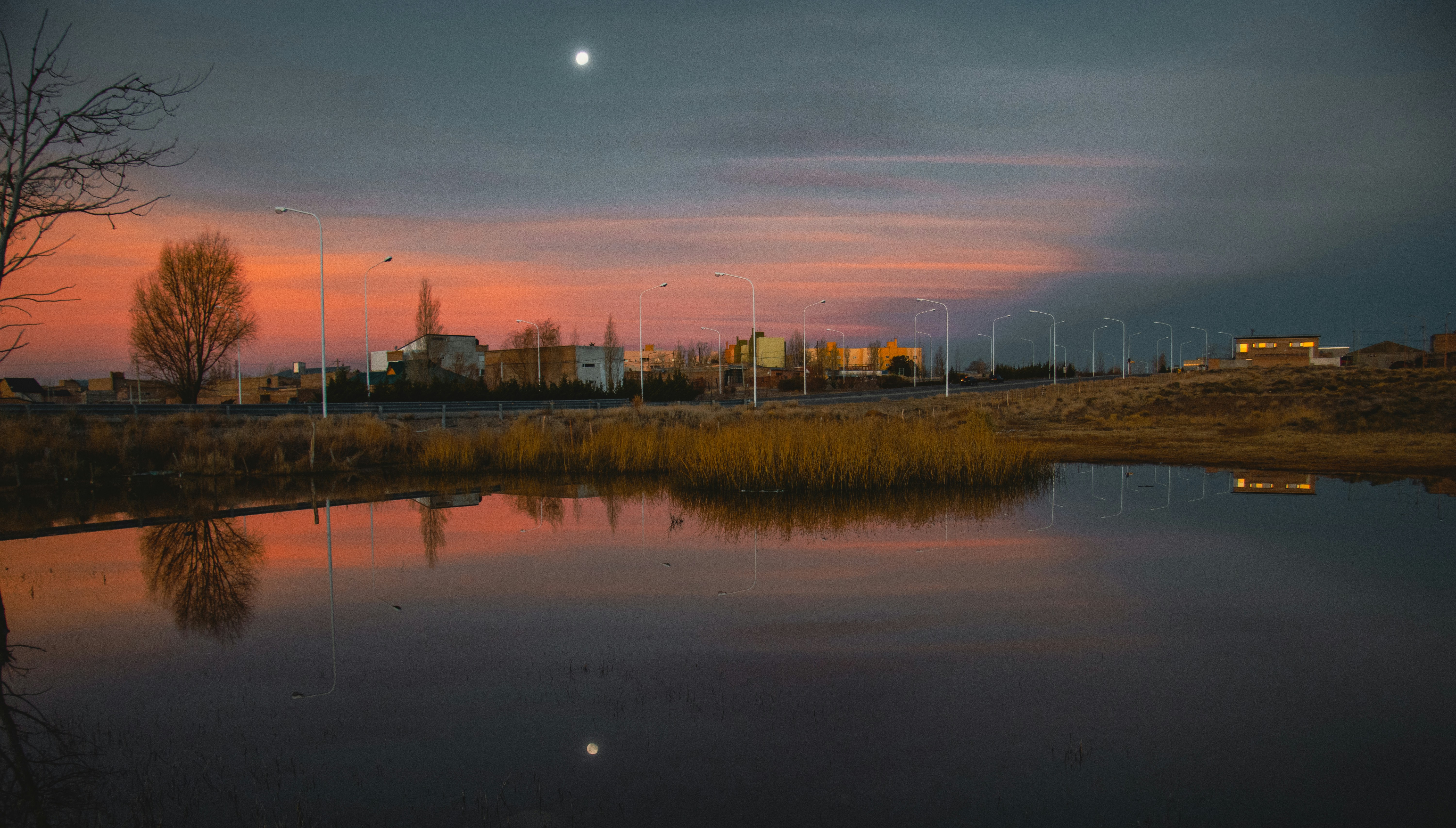 A serene landscape featuring a calm pond reflecting the colorful sky at dusk, with silhouetted trees and distant buildings. The moon hangs prominently above, adding to the tranquil atmosphere.