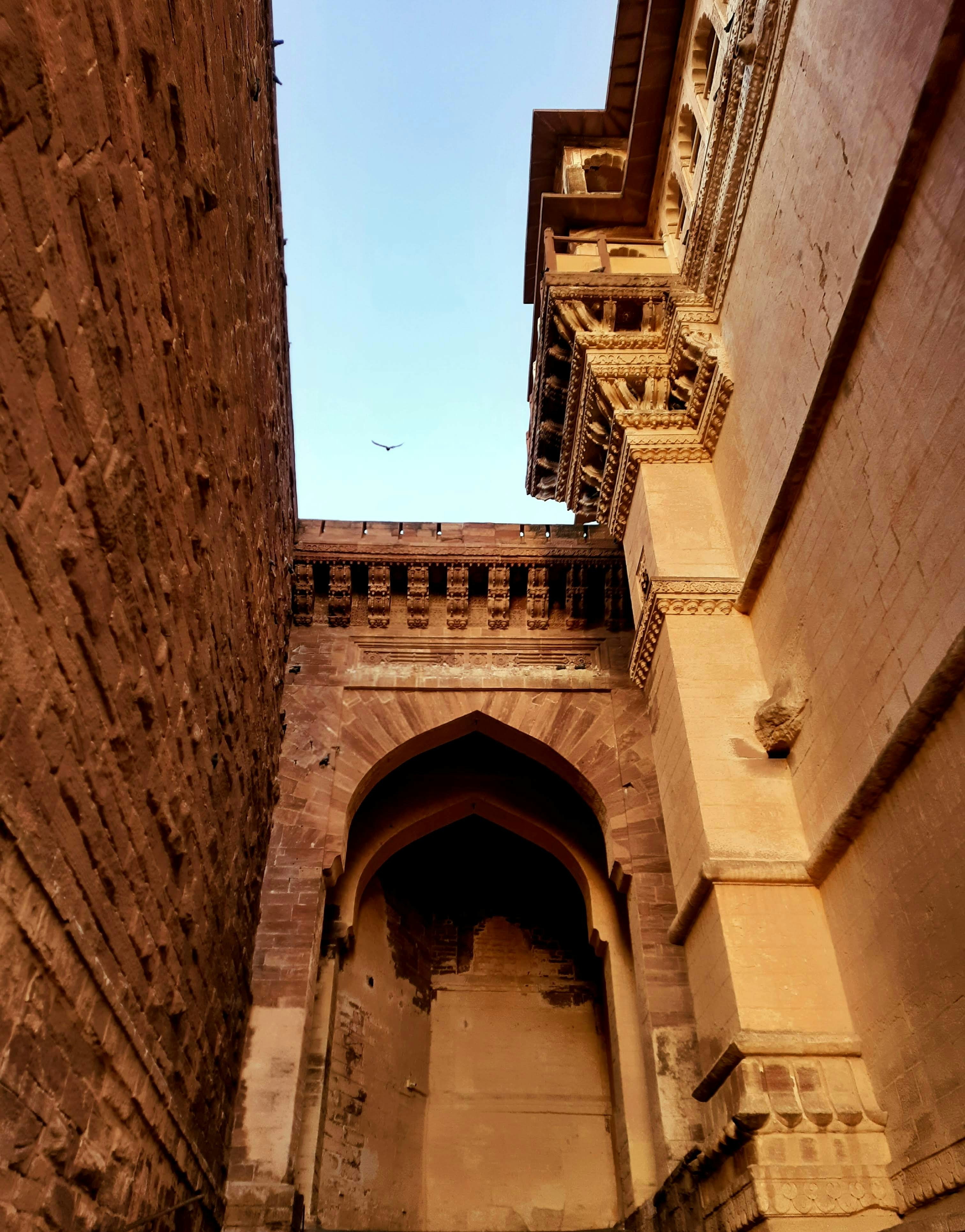 Intricate stonework of a grand archway framed by towering walls, with a clear sky above and a bird in flight.