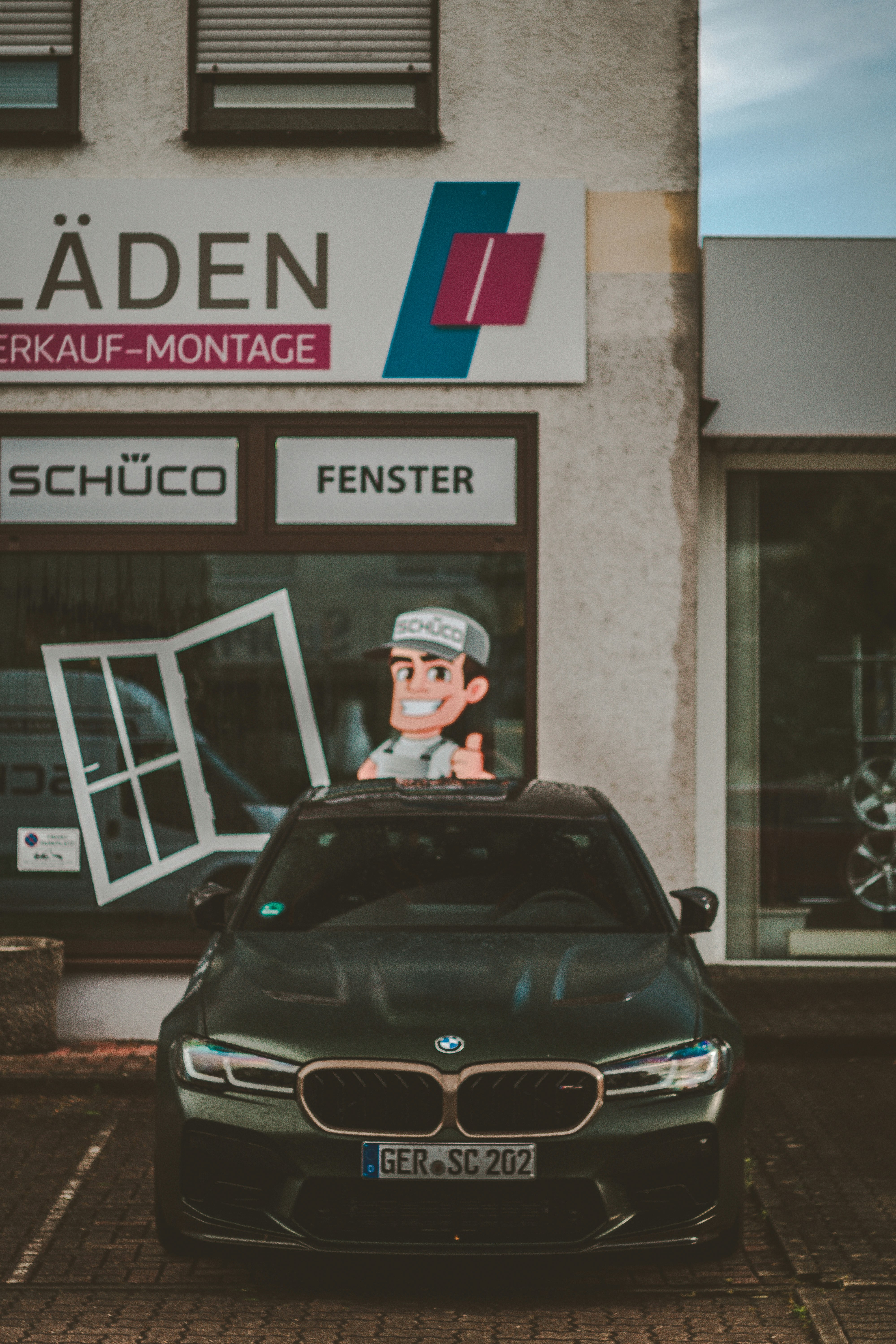 A sleek black BMW parked in front of a window installation shop, featuring a cartoon character promoting the business. The scene captures a blend of modern automotive design and playful advertising.