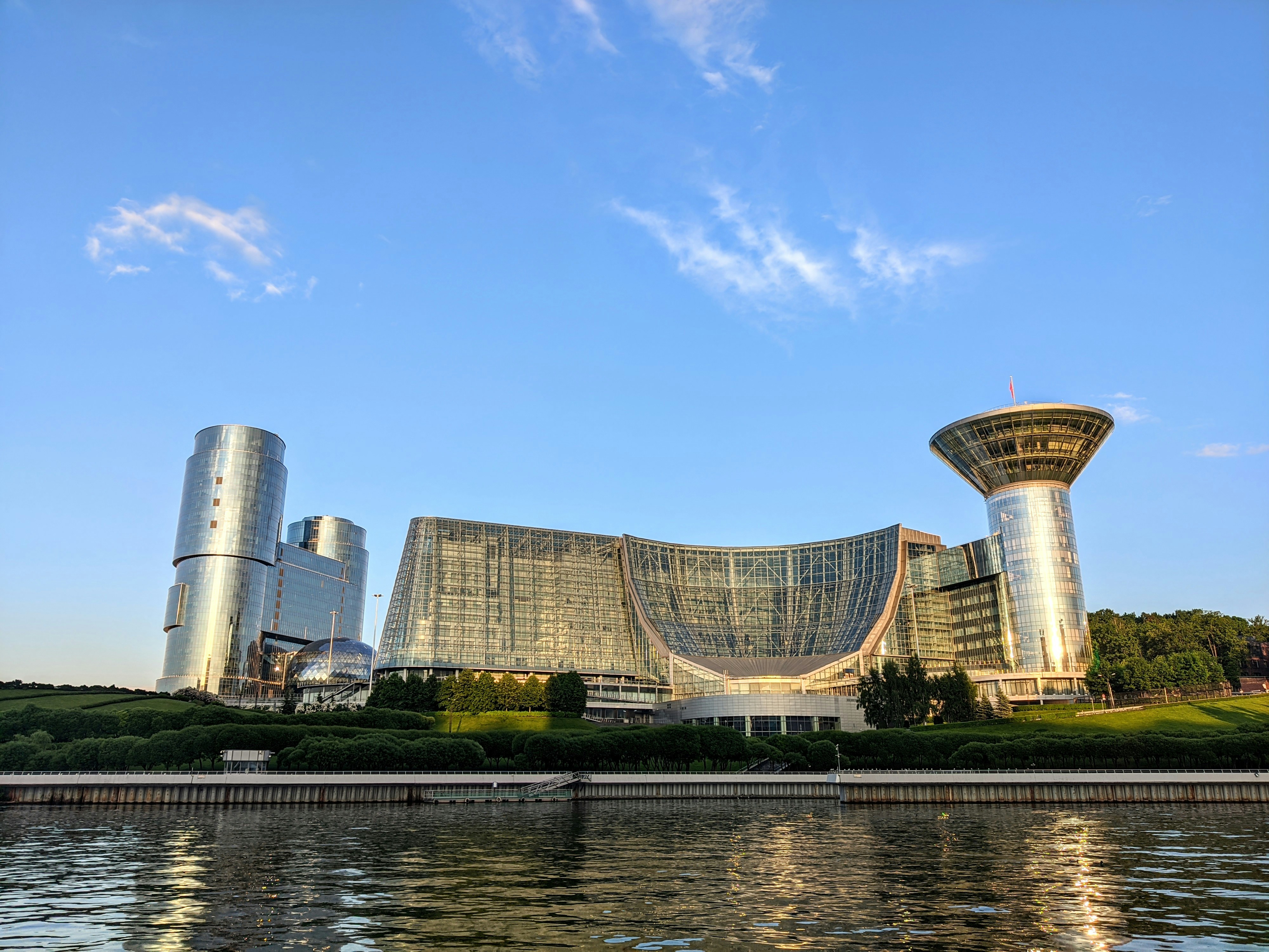 Futuristic building complex with curved glass facades reflecting sunlight, set against a clear blue sky and bordered by a calm water body.