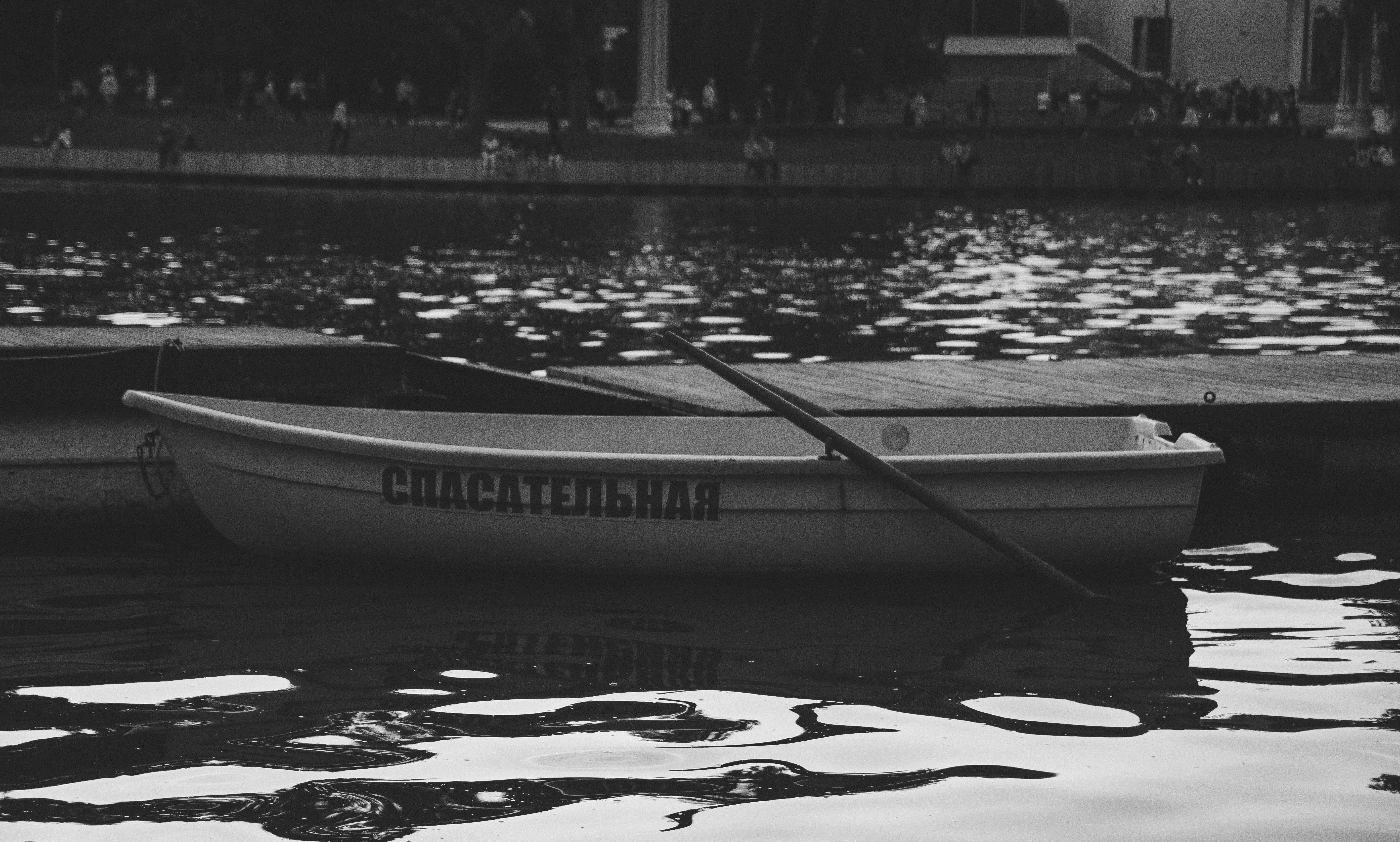 A white rescue boat rests quietly on a dock, its reflection shimmering on the water's surface, surrounded by distant figures enjoying a serene day.
