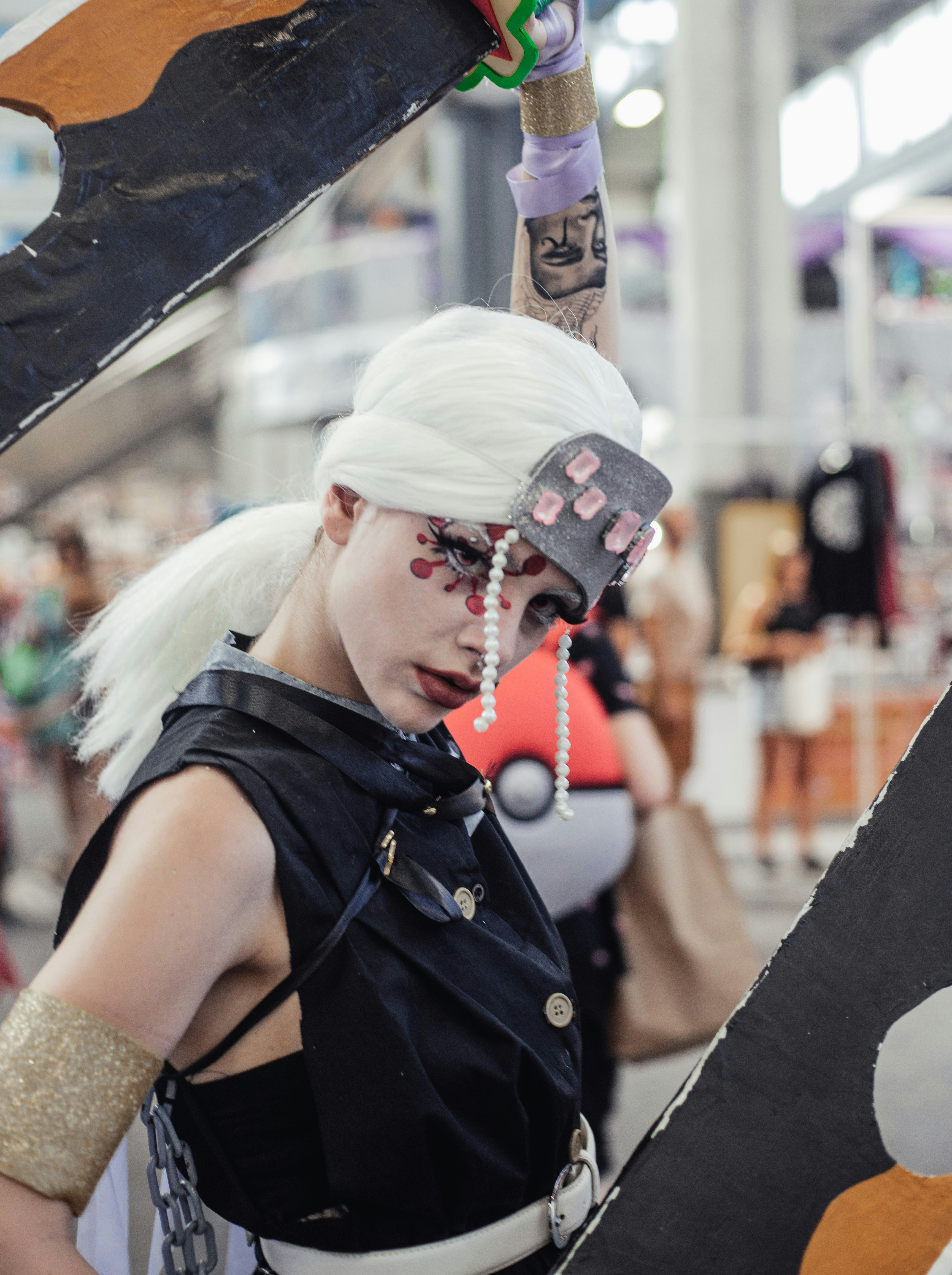 Cosplayer showcasing intricate makeup and costume details, framed by oversized props in a vibrant convention setting.