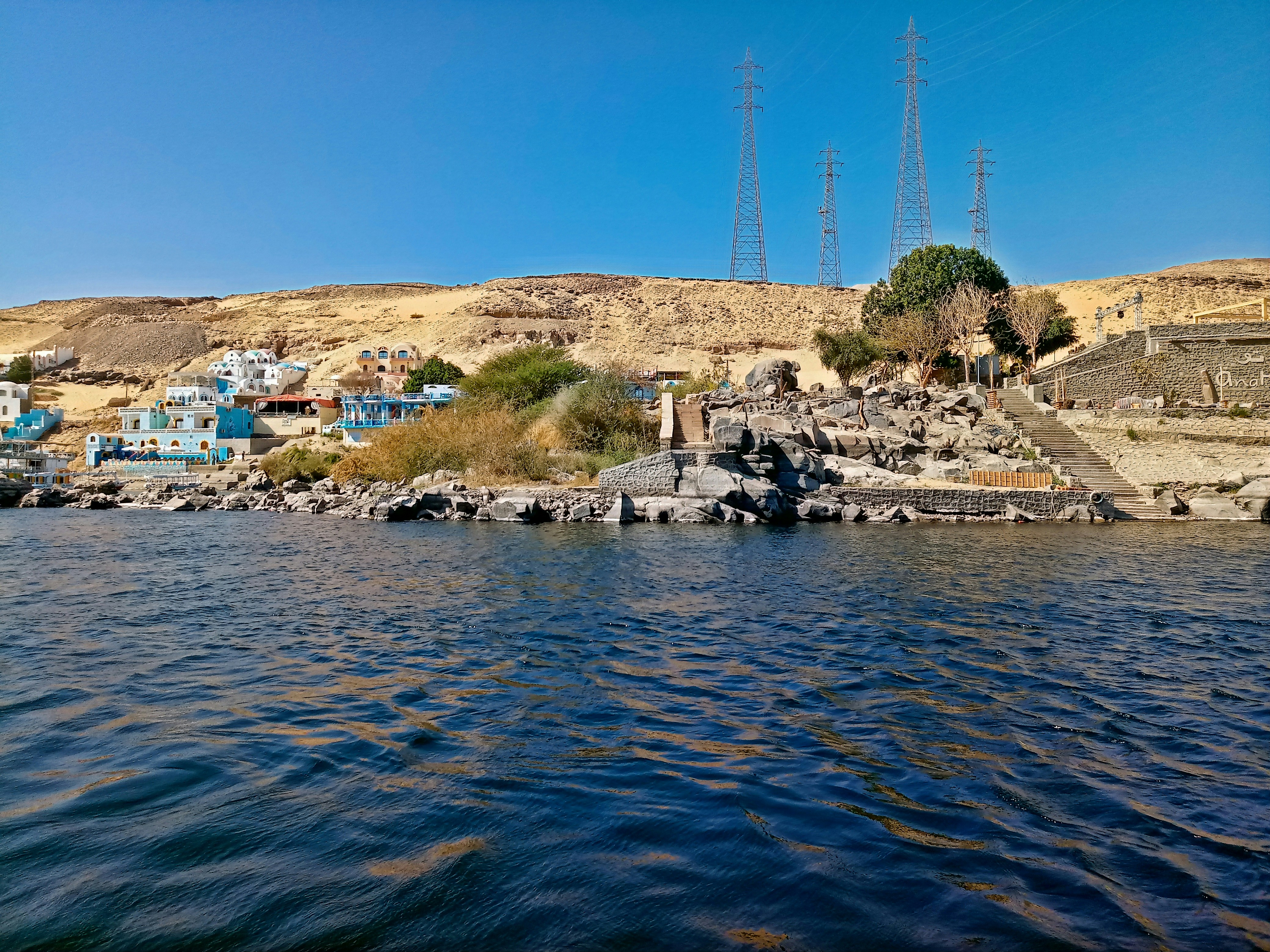 Colorful houses line the rocky shore of a tranquil lake, framed by hills and power lines under a clear blue sky.