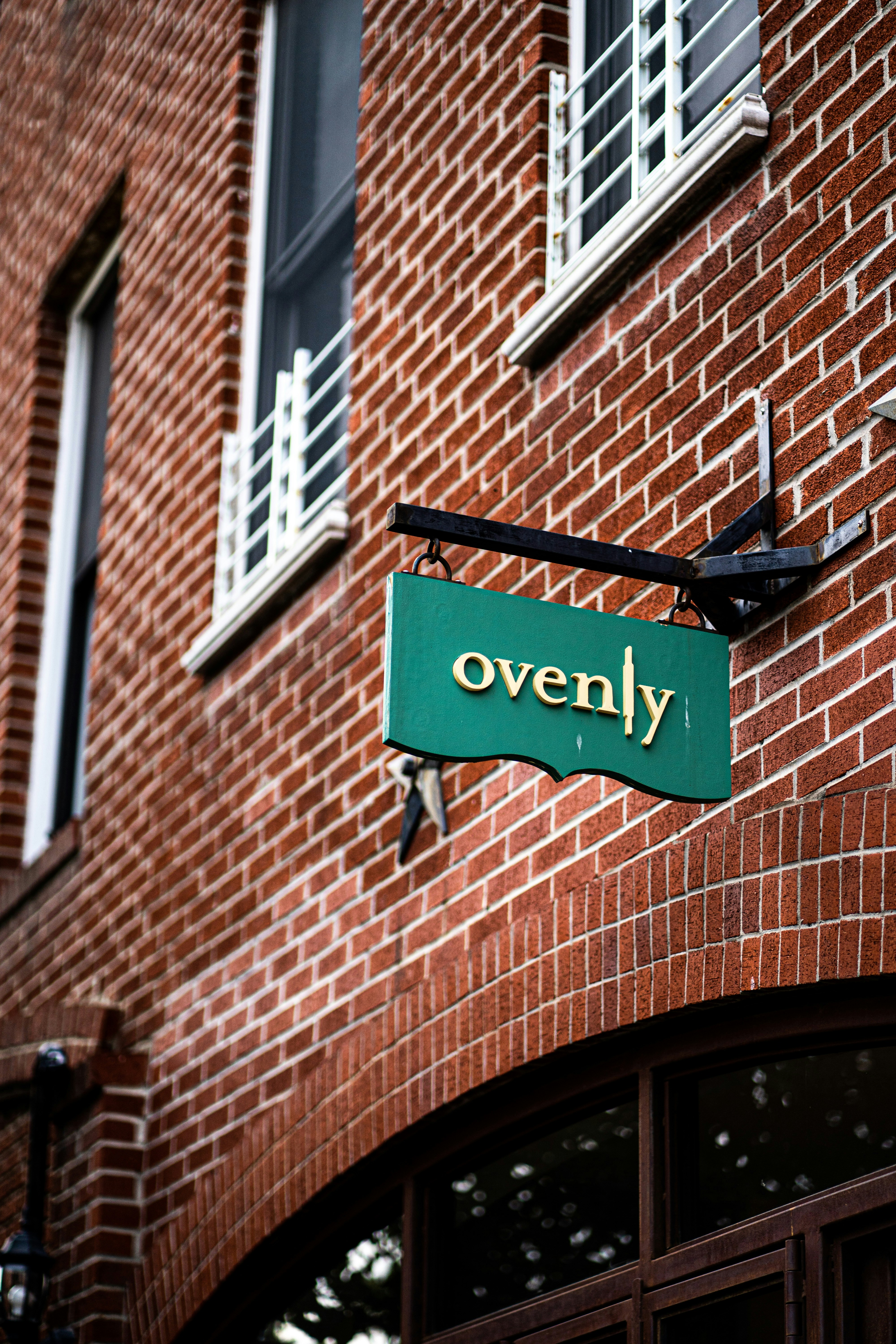 A green storefront sign reading 'ovenly' hangs from a black bracket on a red brick wall above an arched storefront window.