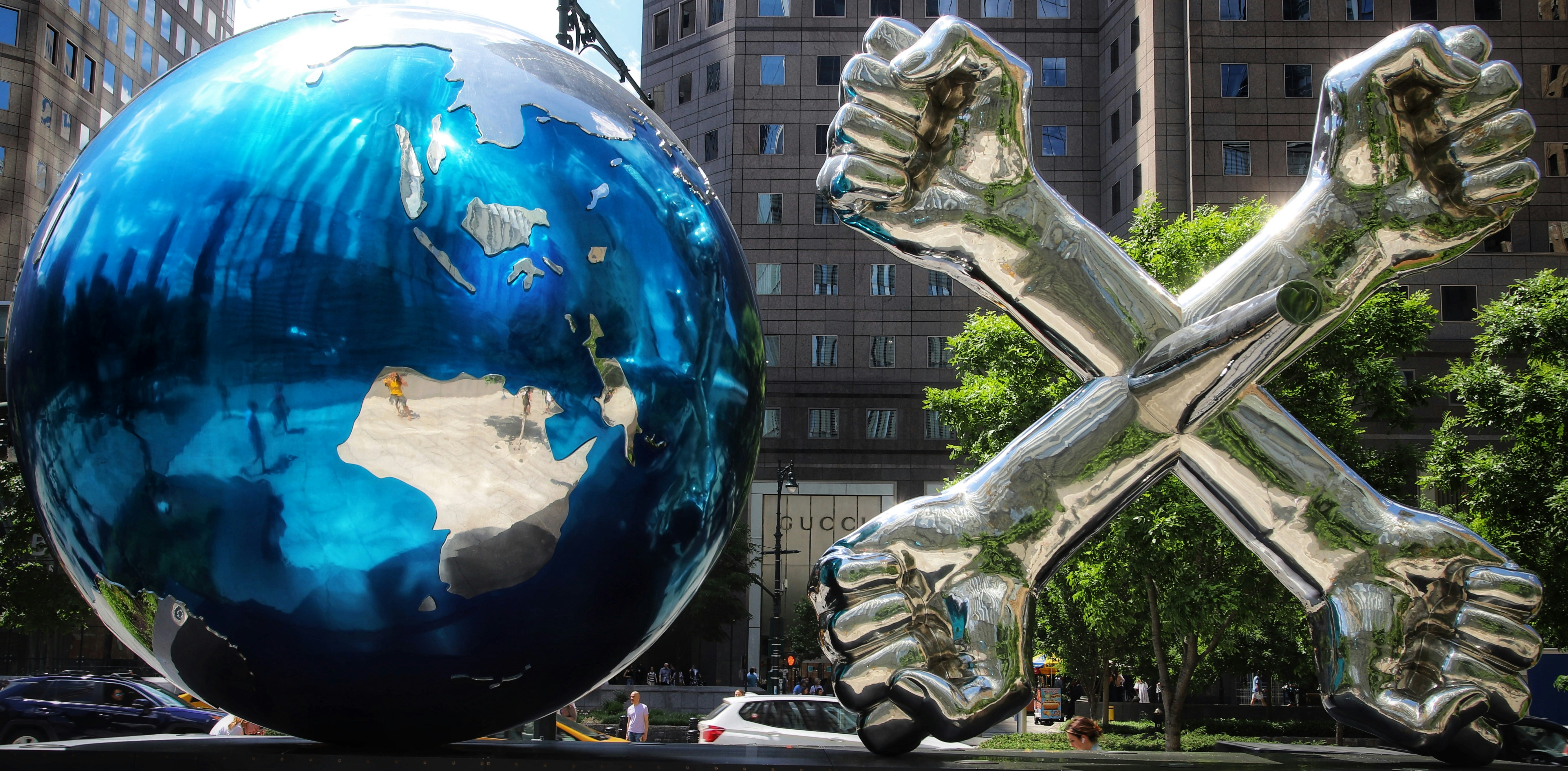 Sculpture of a blue globe alongside metallic crossed arms in an urban setting.