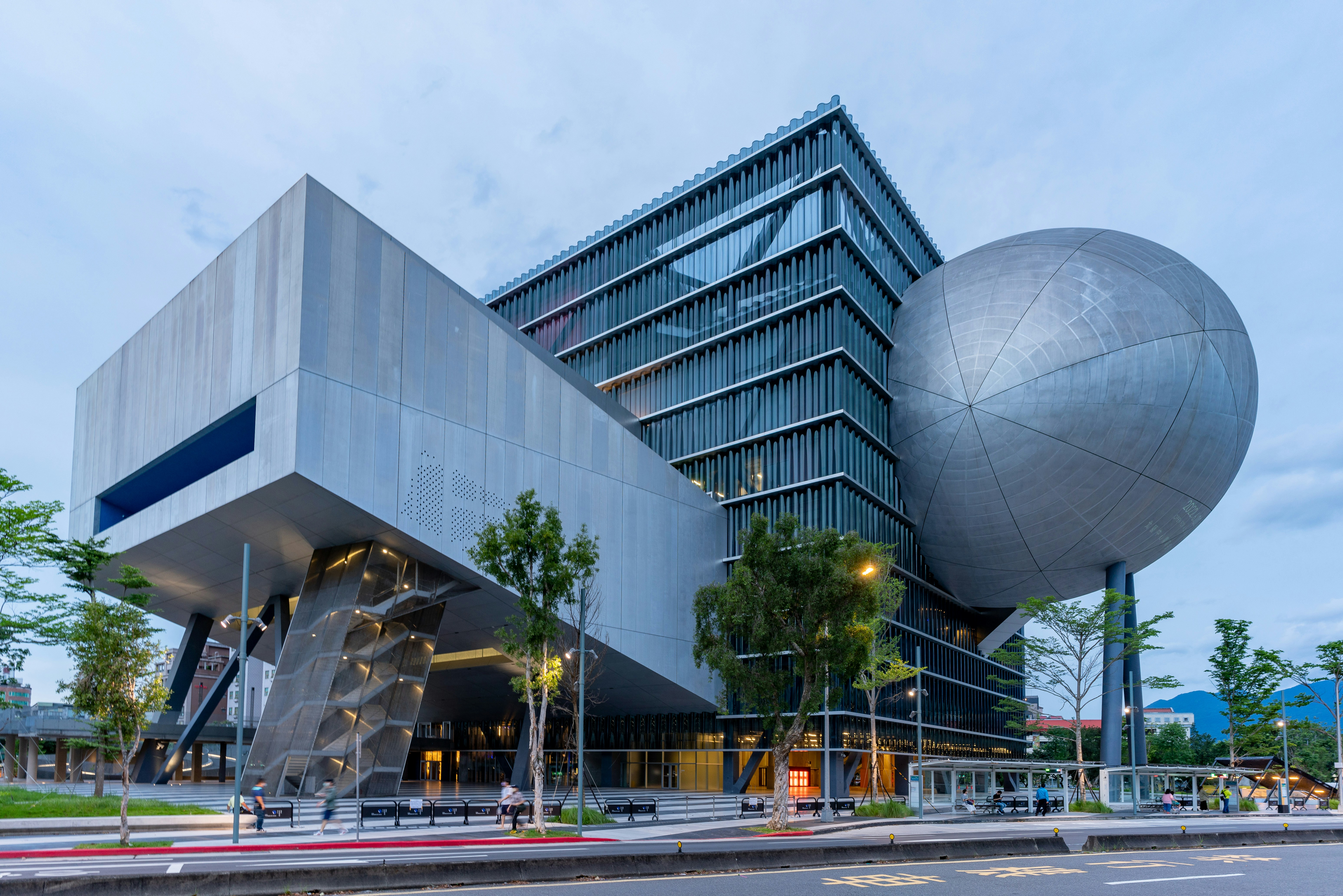 Modern building featuring a striking spherical structure and angular design elements, surrounded by greenery and urban landscape.