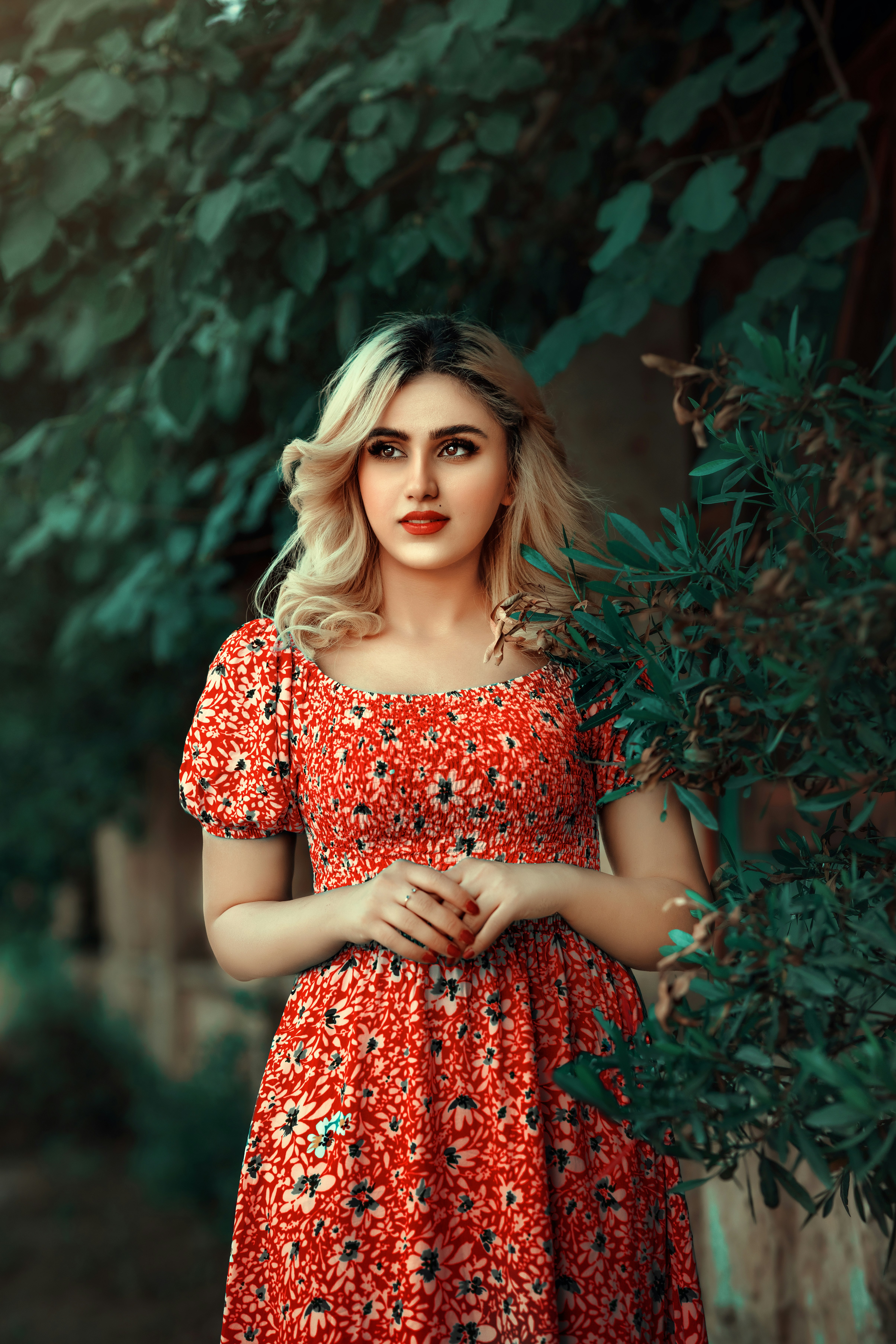 Young woman in a vibrant floral dress stands among lush greenery, exuding a serene yet confident presence. Her gaze reflects a moment of contemplation.
